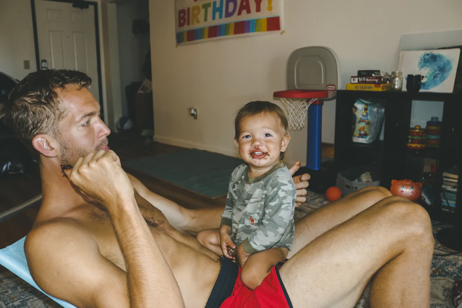 A father relaxes on a couch with his smiling toddler, both enjoying a playful moment. The room is filled with colorful toys and decorations.