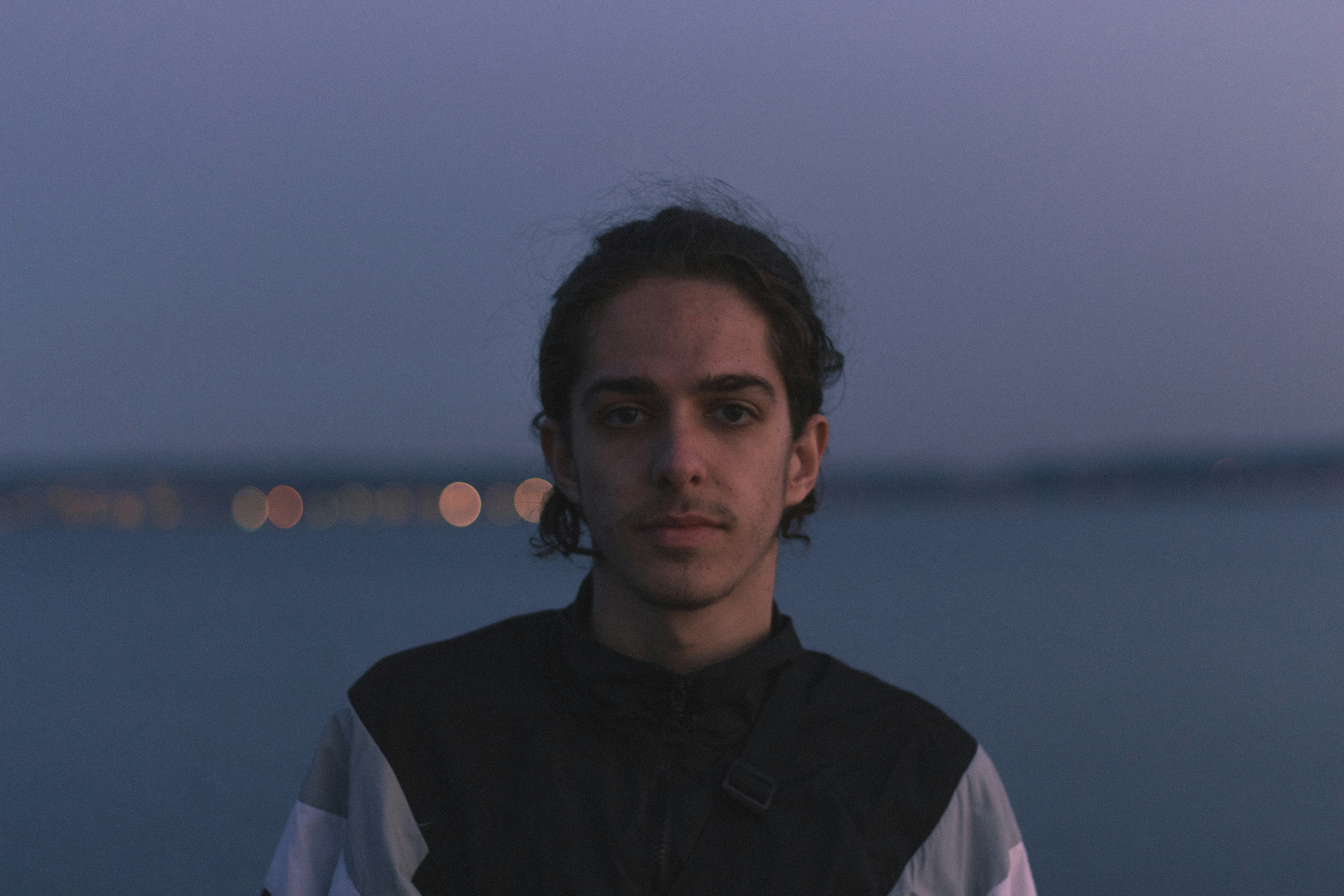 Young man standing by the water's edge during twilight, with soft bokeh lights in the background. The calm atmosphere enhances the contemplative mood.