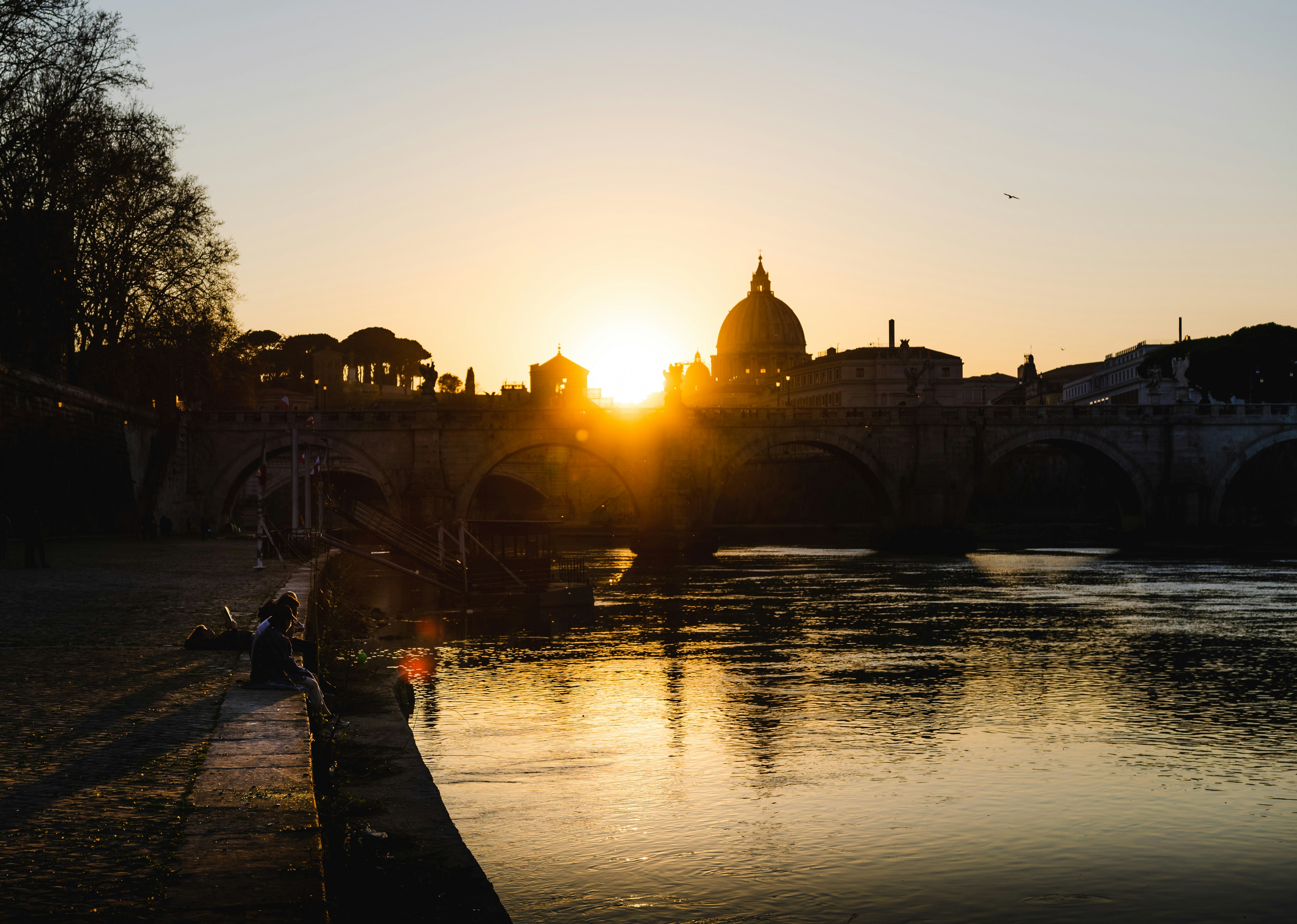 Silhouette of a bridge and cityscape against a setting sun reflected in a tranquil river.