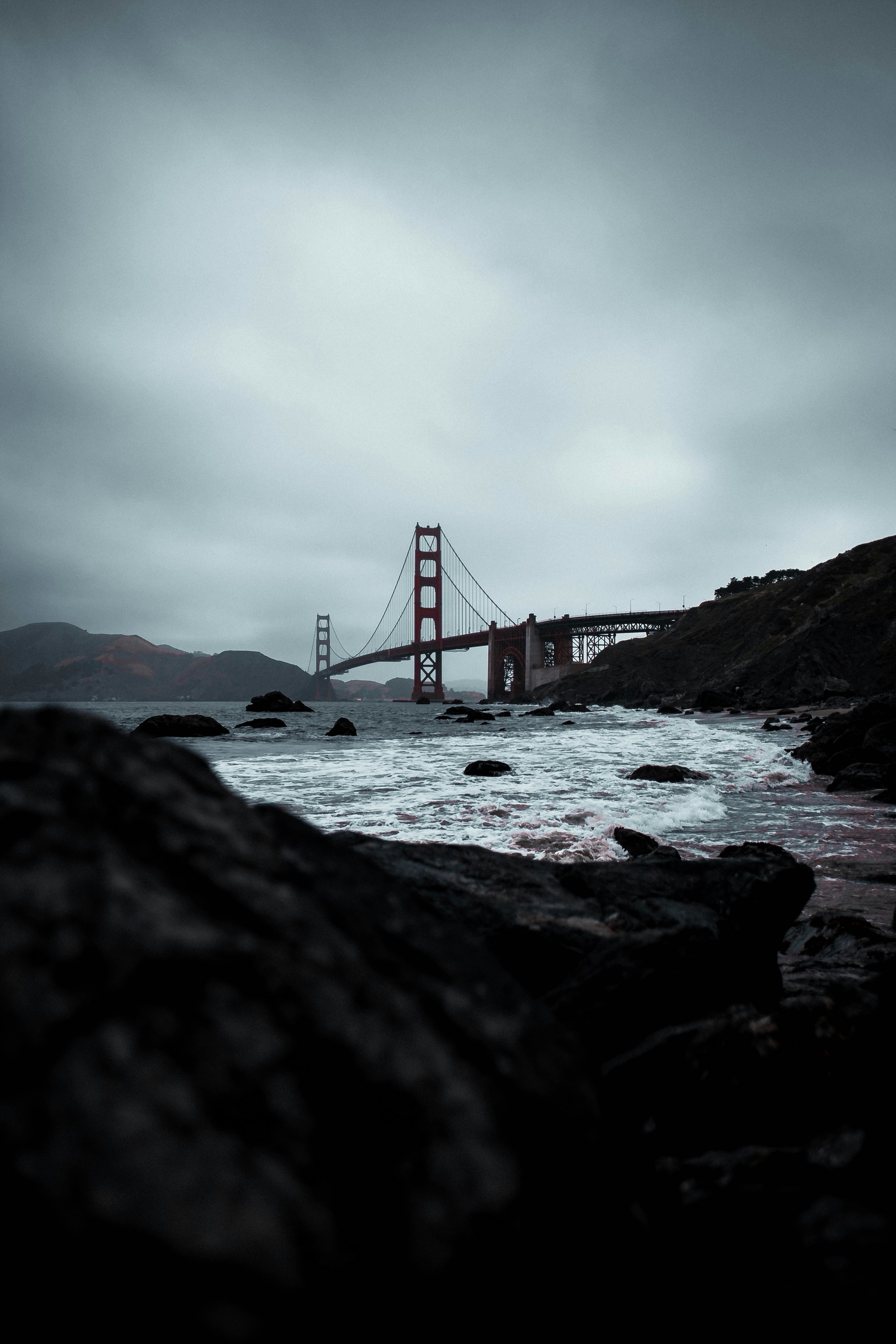 Golden Gate Bridge shrouded in moody clouds, viewed from rocky shoreline with gentle waves lapping at the stones.
