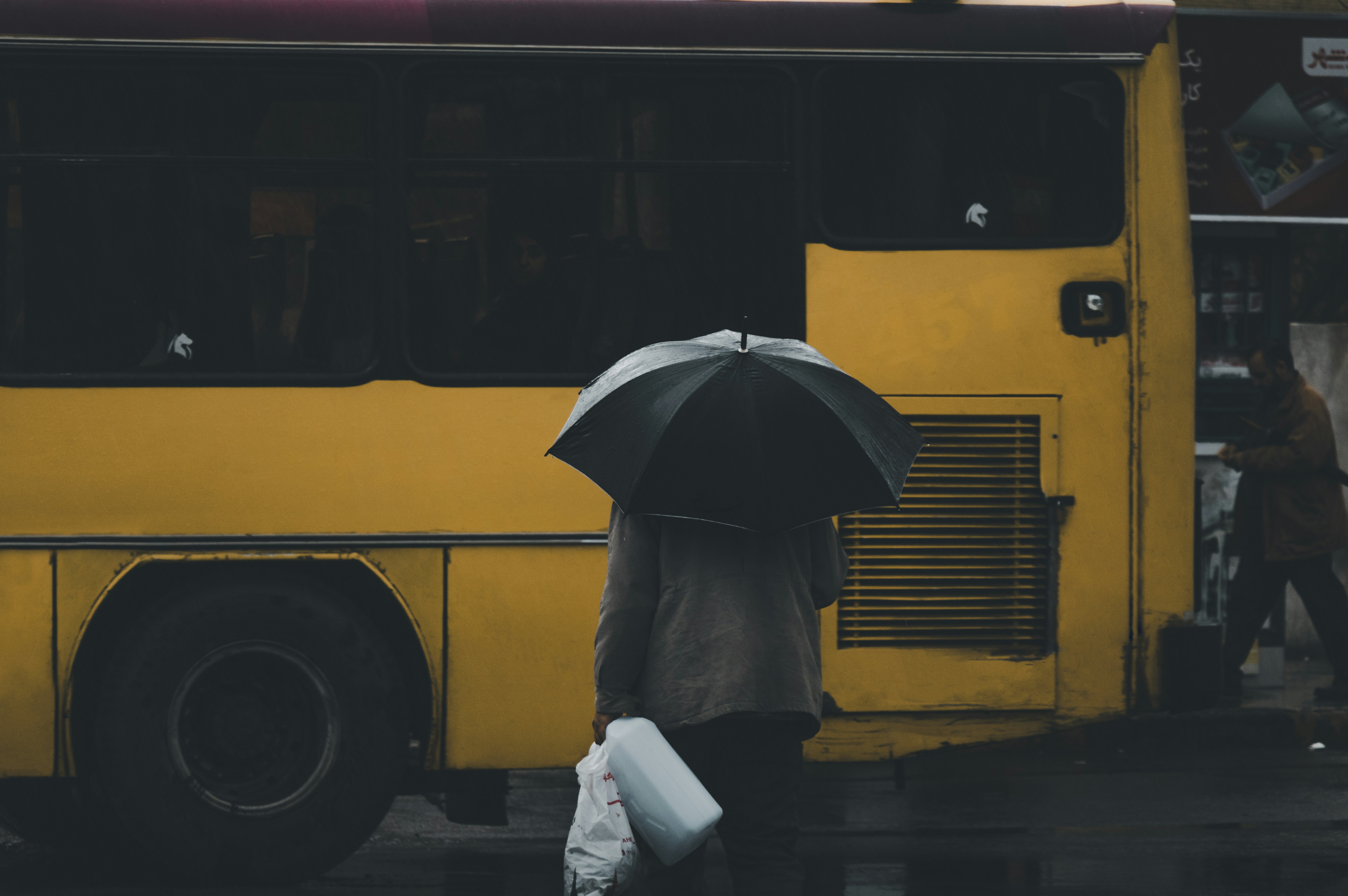 A figure with an umbrella walks beside a vibrant yellow bus in a rainy urban setting.