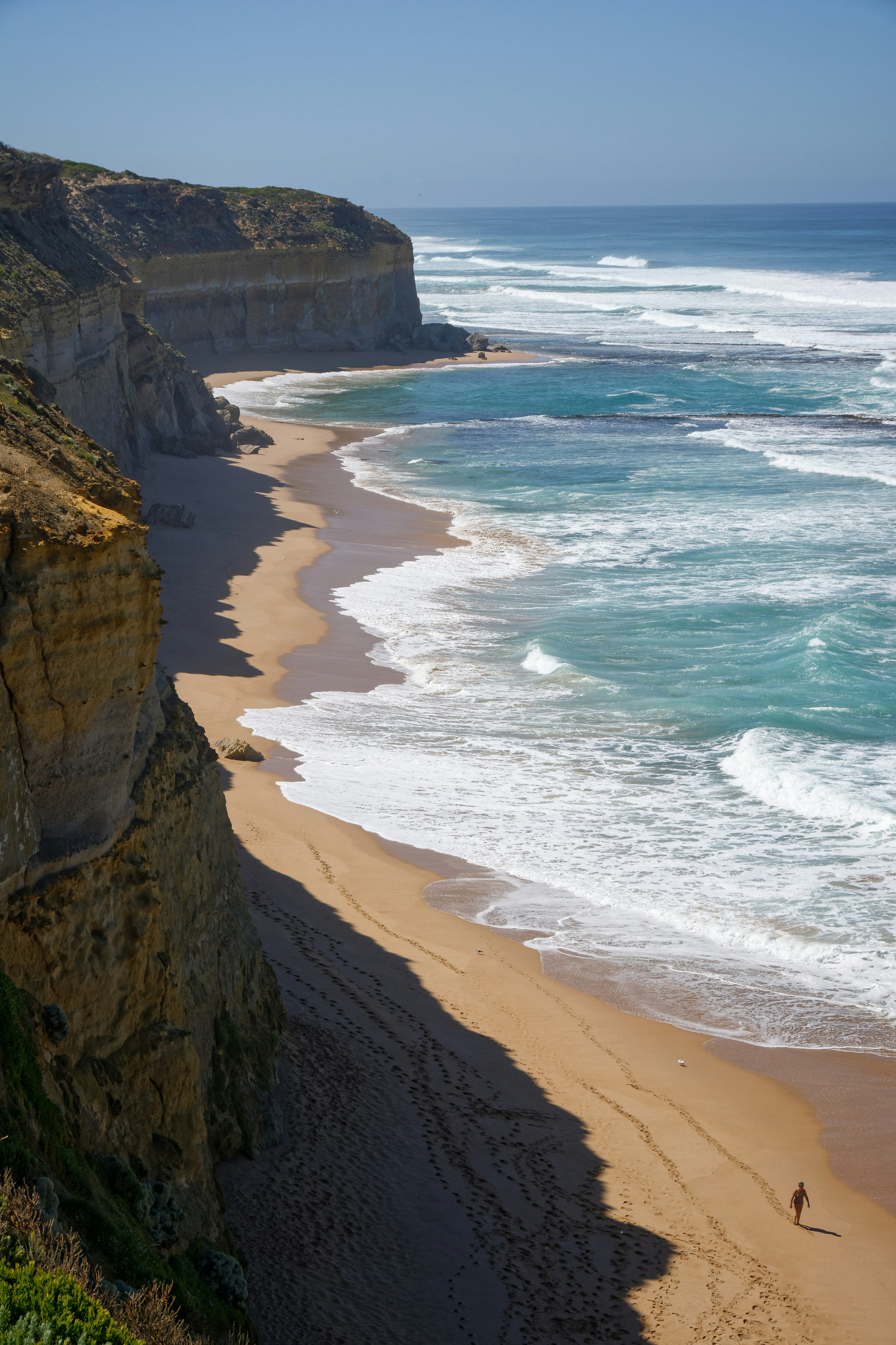 A solitary figure walks along a sandy beach bordered by towering cliffs, with waves gently lapping at the shore under a clear blue sky.