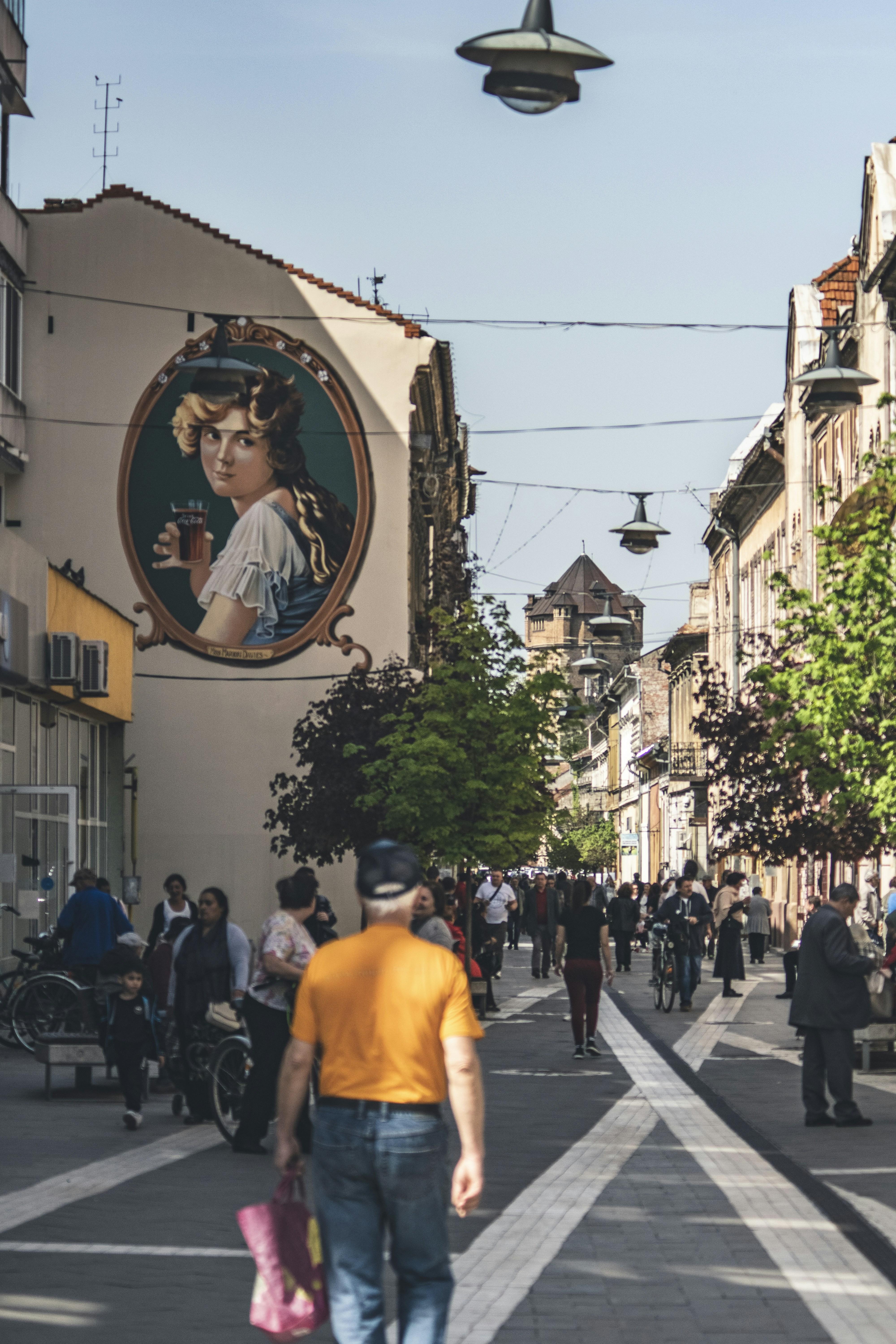 A vibrant street scene featuring a mural of a girl holding a drink, surrounded by pedestrians and greenery, capturing the essence of urban life.