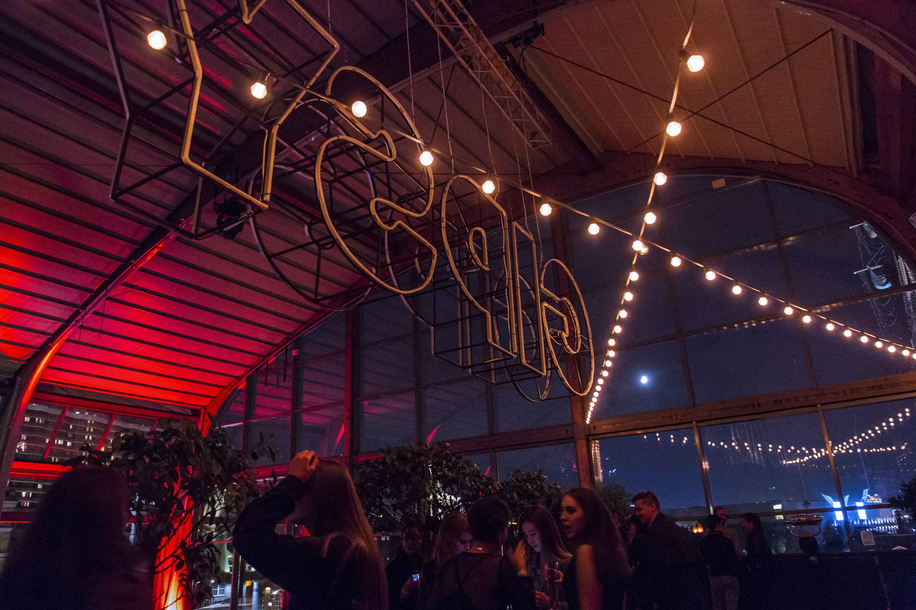 People socializing beneath a neon sign and string lights in a glass-roofed venue at night.
