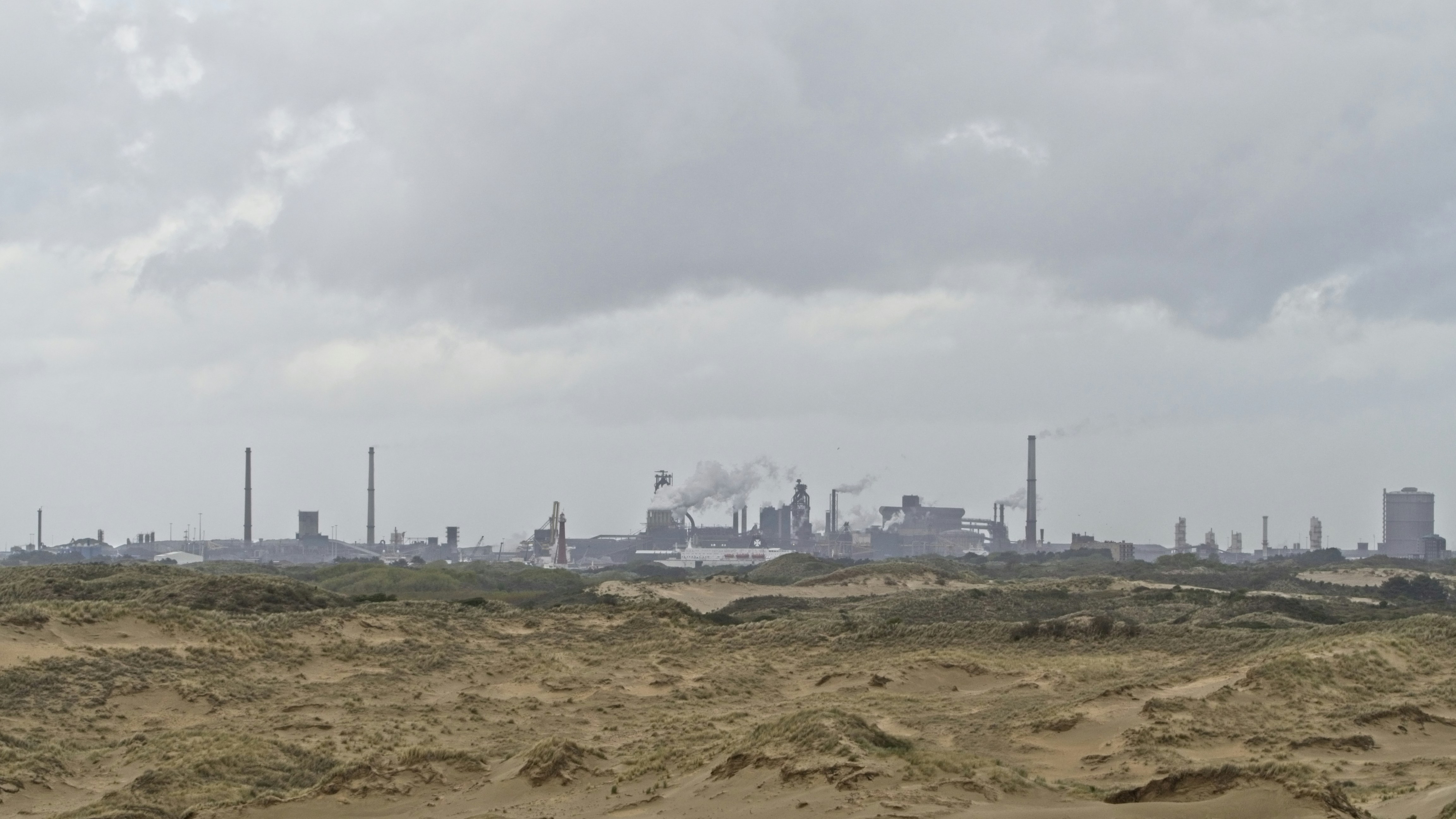 A distant industrial skyline looms over a sandy landscape, with smoke rising from chimneys under a cloudy sky.
