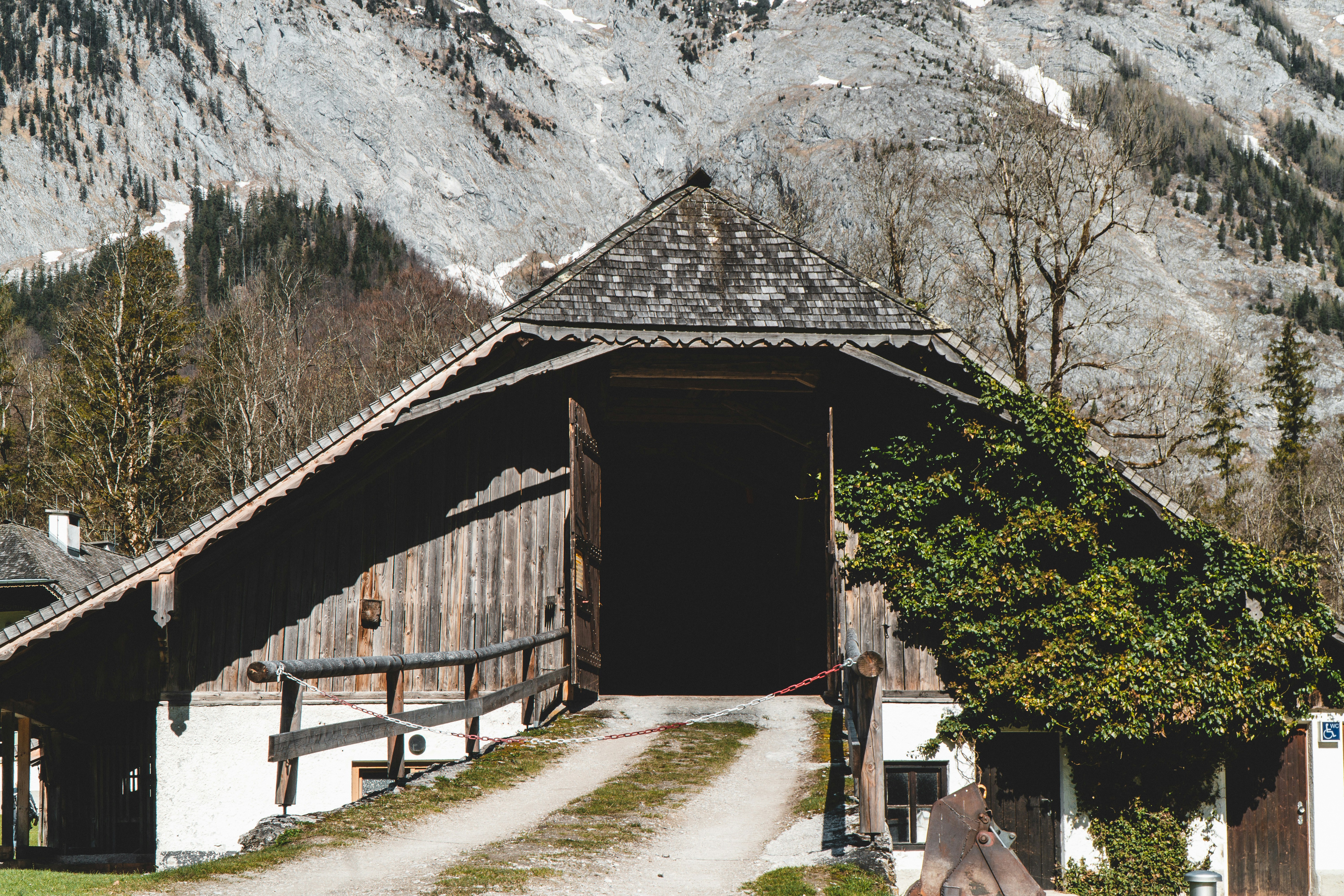 A traditional wooden barn with an open entrance, framed by lush greenery and the majestic mountains in the background.