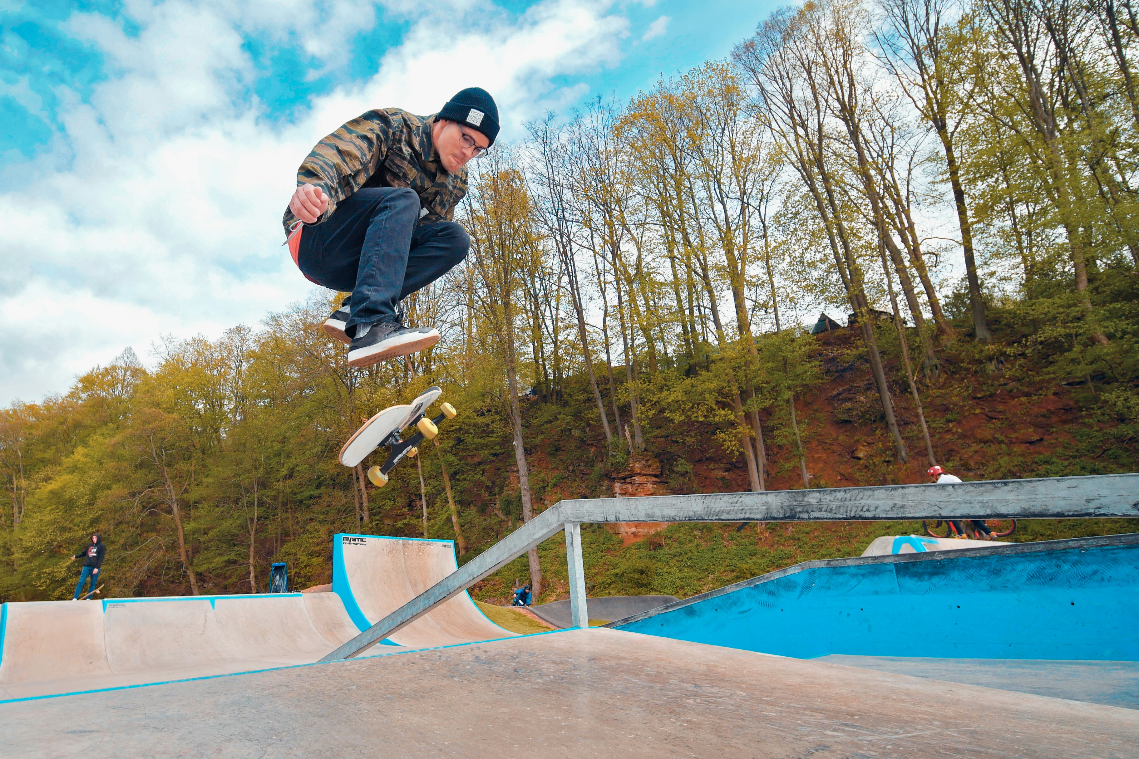 Skateboarder executing a pop shove-it above a ramp in a vibrant skate park surrounded by trees.