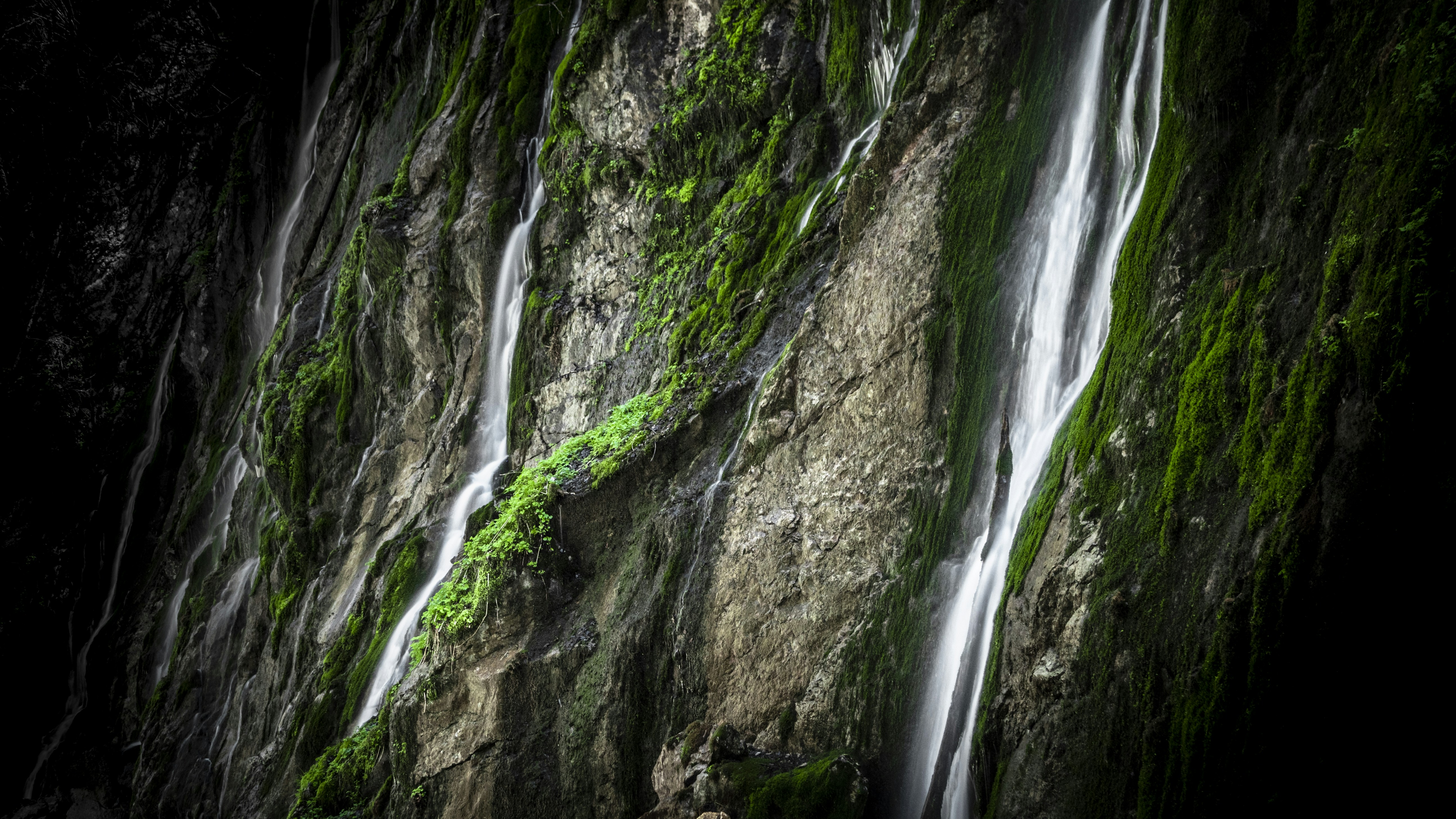 Thin waterfalls cascade down a moss-covered rocky cliff, creating a natural tapestry of green and gray.