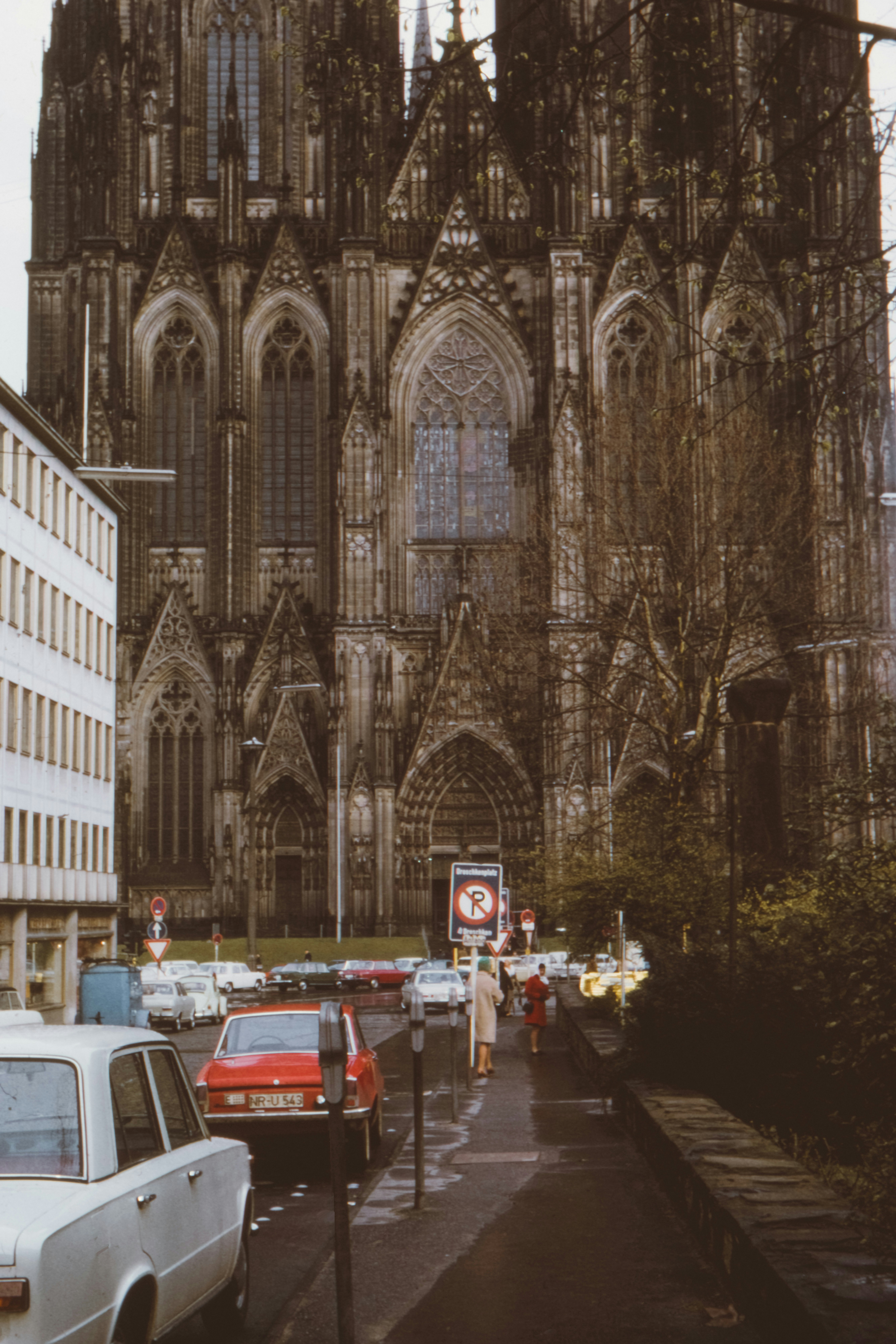 A Gothic cathedral towers over a street lined with parked cars and pedestrians.