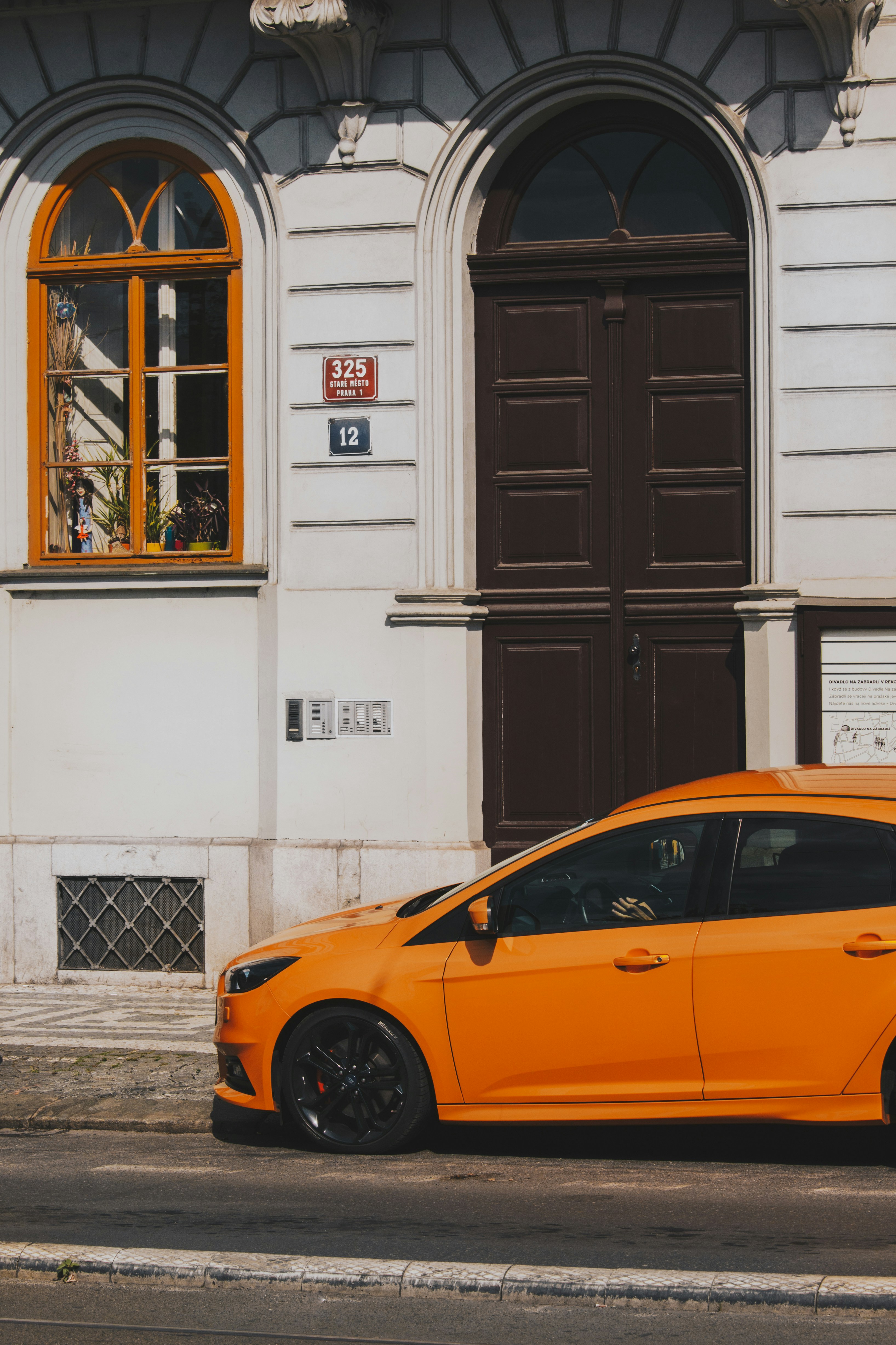 Bright orange car parked beside a classic building with ornate architecture and contrasting window frames.