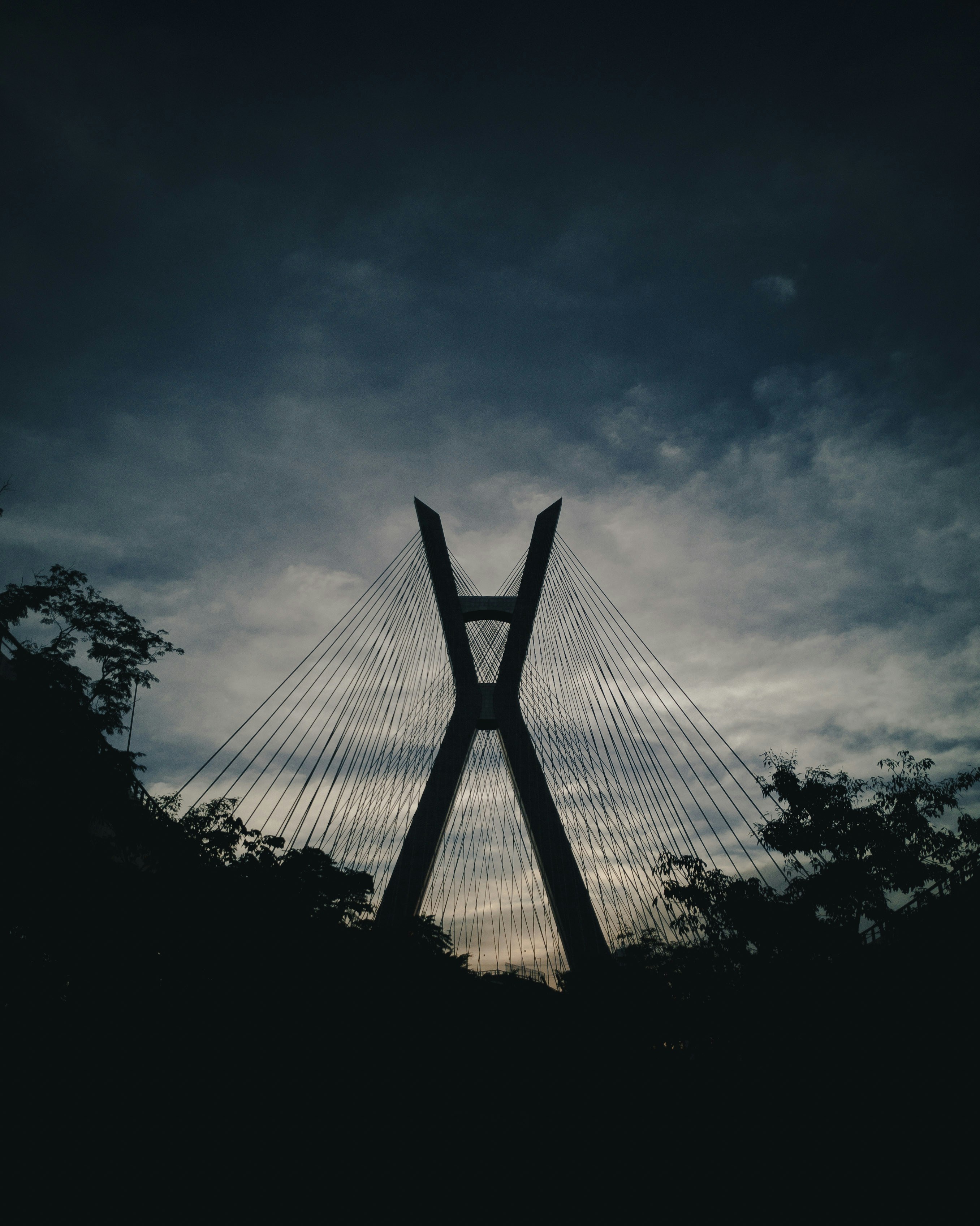 Silhouette of a modern suspension bridge framed by a dramatic twilight sky, showcasing intricate cable patterns and structural design.