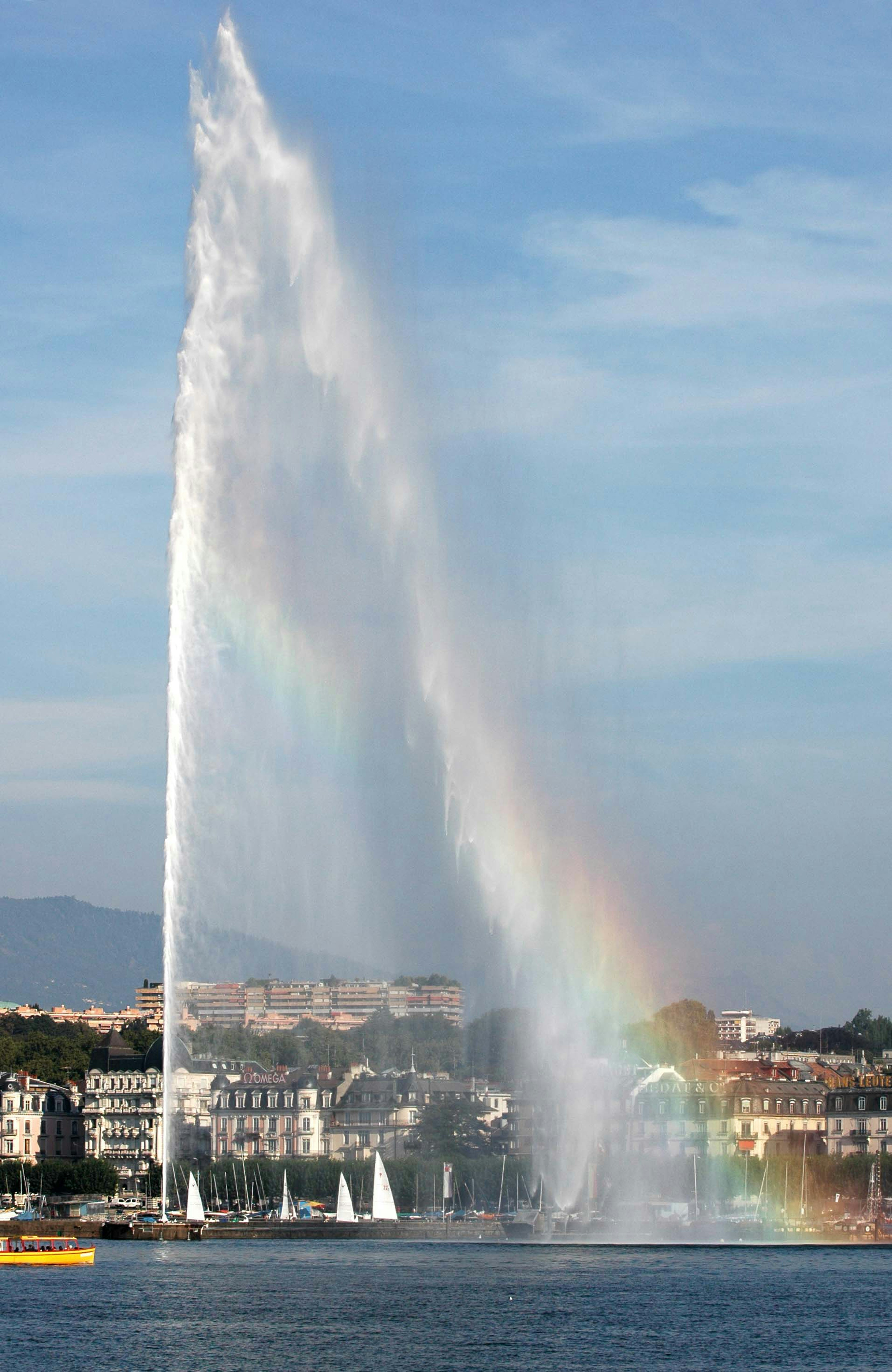 Jet d'Eau fountain in Geneva creating a vibrant rainbow against a clear blue sky, with sailboats dotting the harbor below.