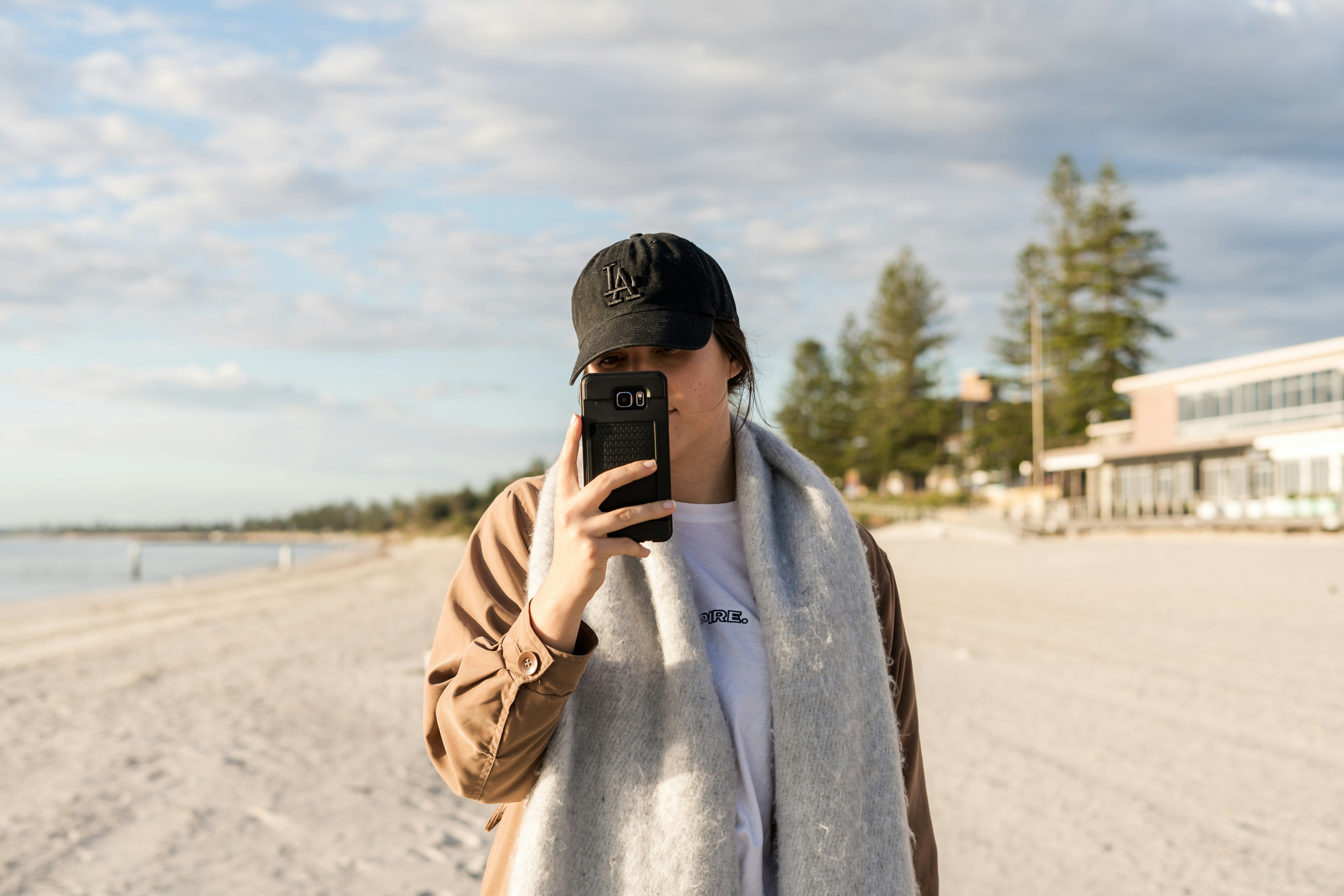 Individual holding a smartphone while standing on a sandy beach, with a backdrop of trees and a coastal building. Soft light enhances the scene's tranquility.