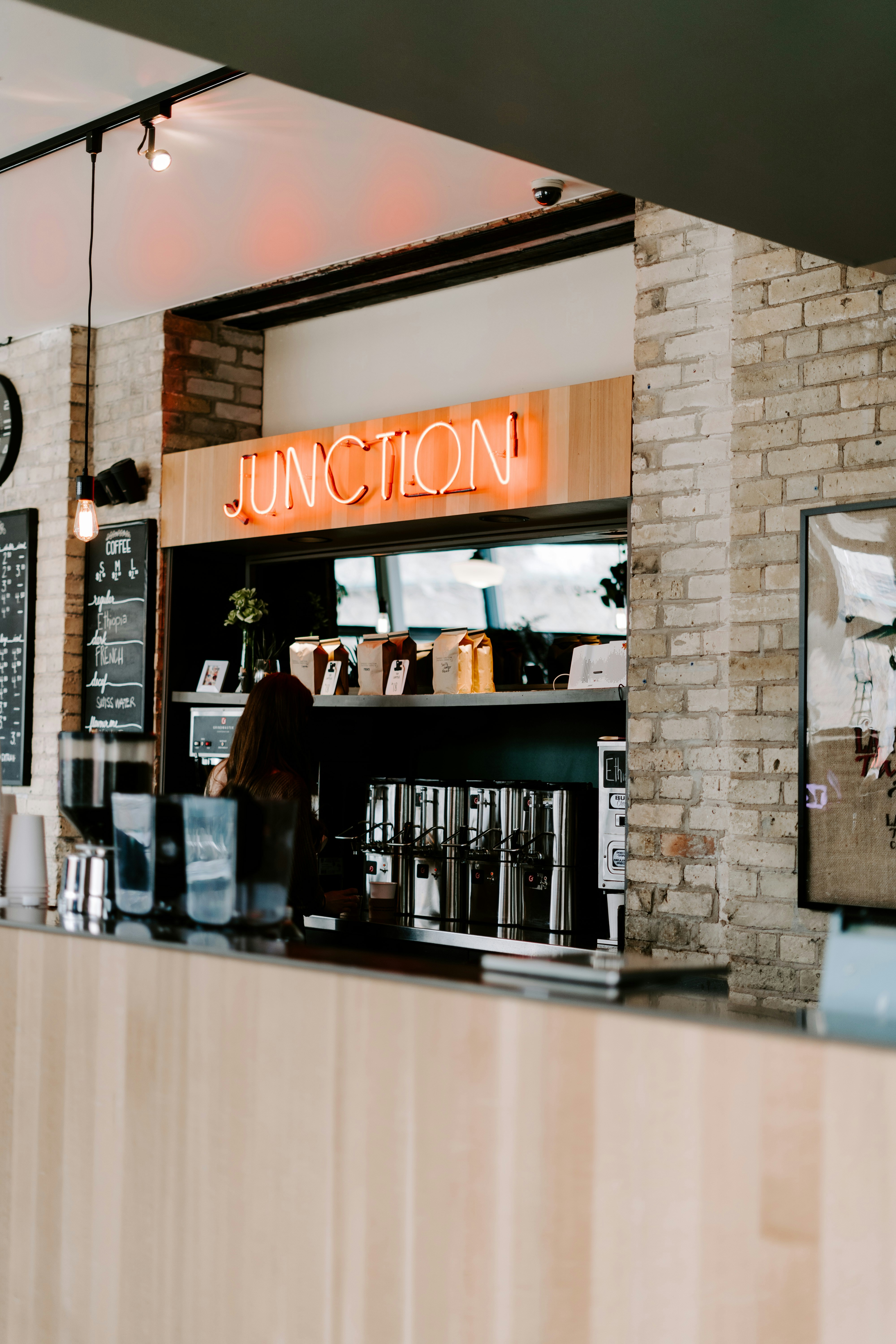 Neon 'JUNCTION' sign above a brick café counter, with equipment and a barista in the background.