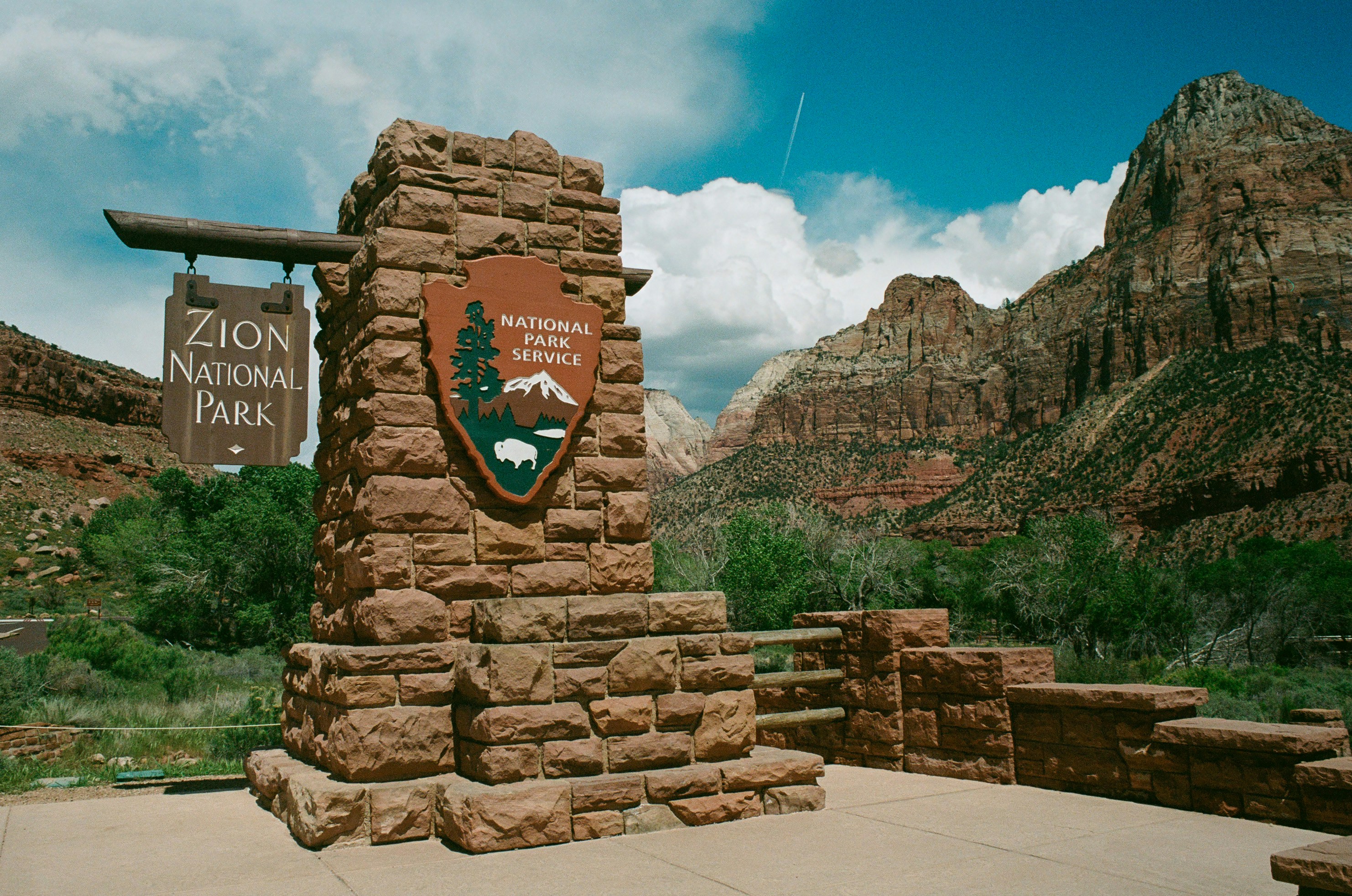Zion National Park entrance sign crafted from stone, surrounded by vibrant landscapes and towering cliffs. The park's emblem showcases its natural beauty.