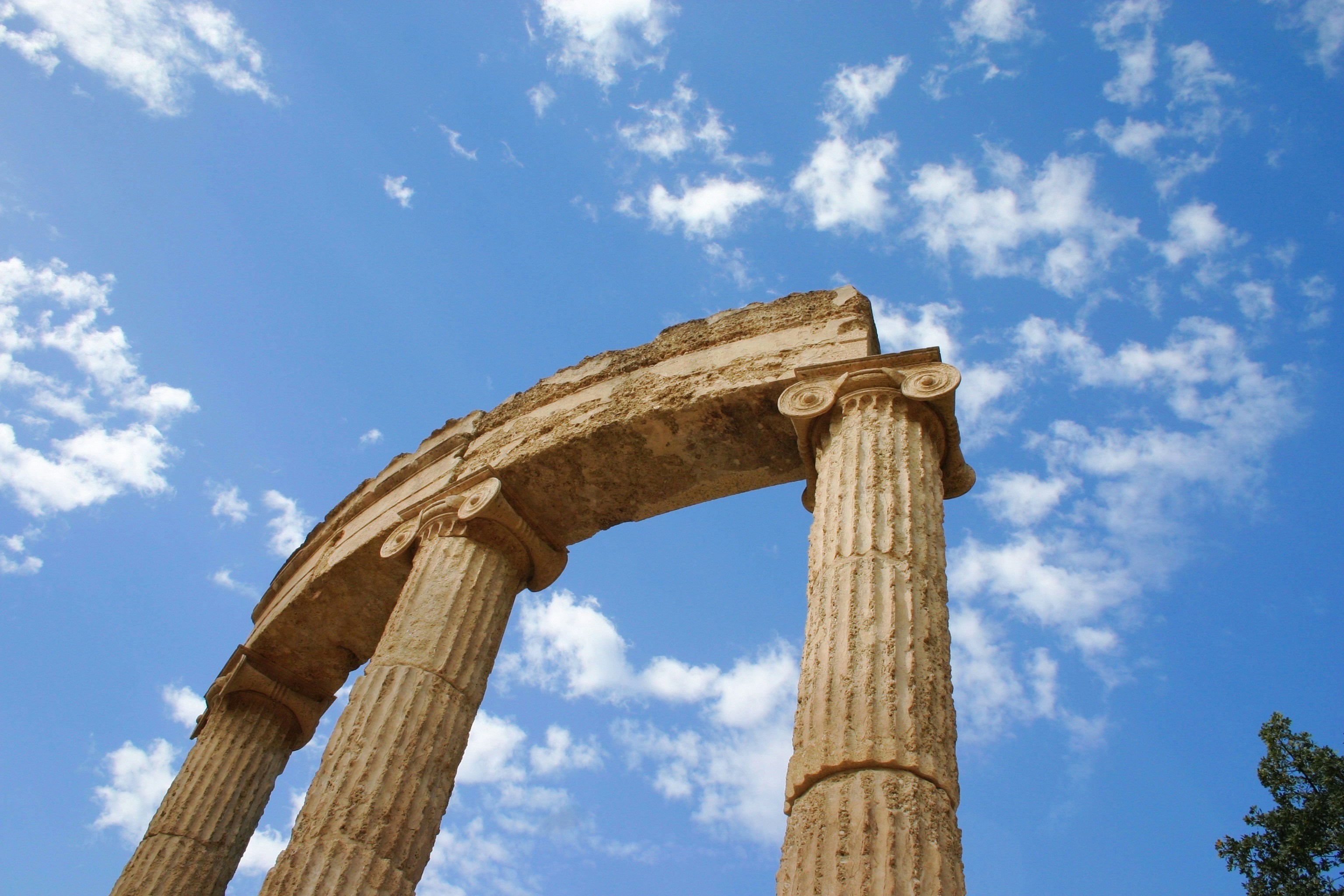 Weathered columns supporting a partially intact arch against a vibrant blue sky with scattered clouds.