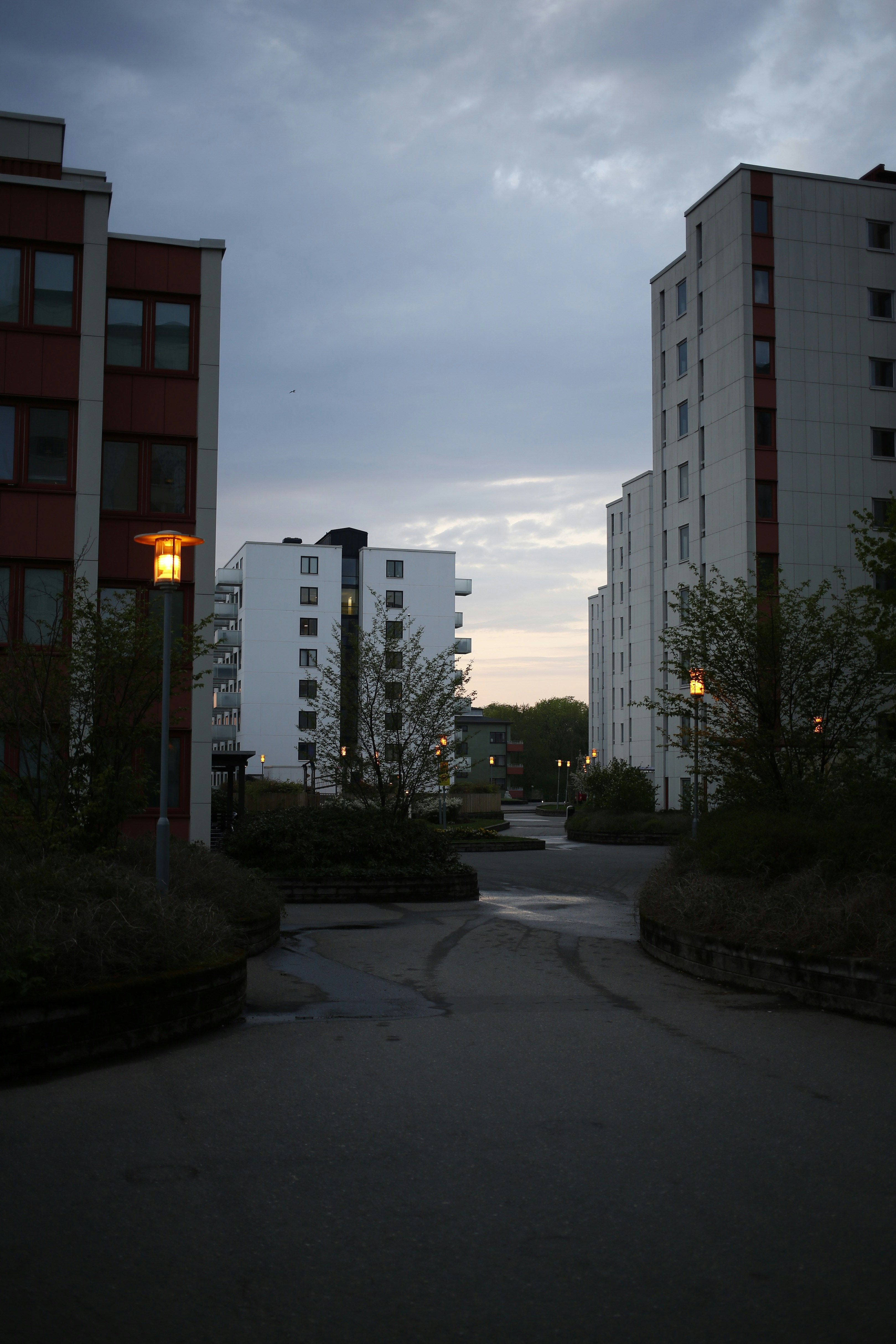 Quiet urban pathway flanked by modern buildings under a cloudy sky, illuminated by street lamps. The scene invites exploration and reflection.
