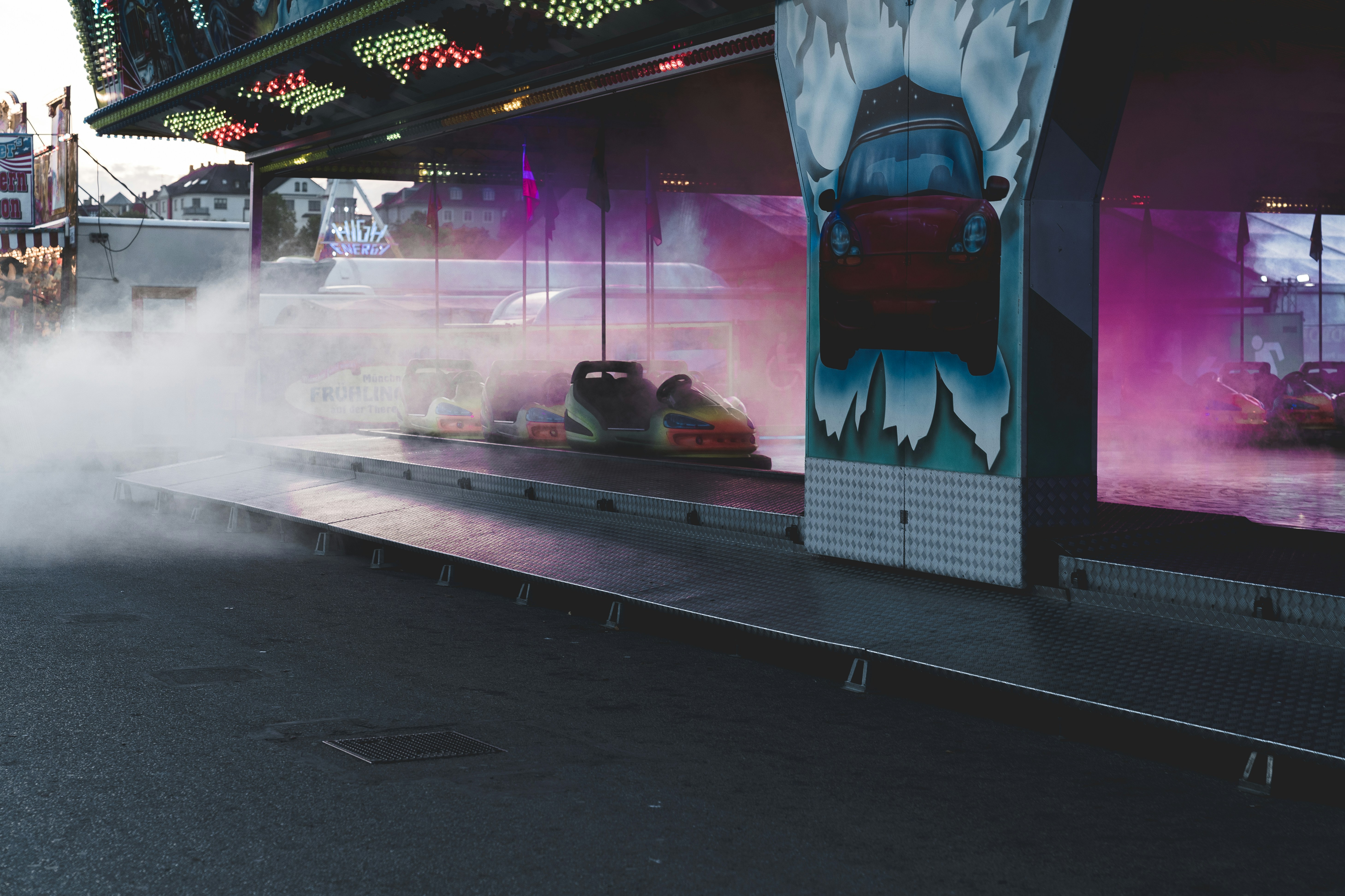 Colorful bumper cars shrouded in mist and illuminated by neon lights at an amusement park attraction.