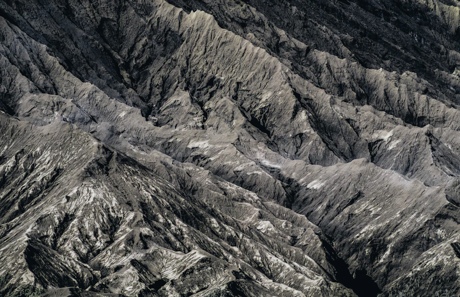 Craggy gray mountain ridges and eroded gullies form a dense, textured landscape under moody light. Dark shadows carve through the slopes, emphasizing the sharp relief.