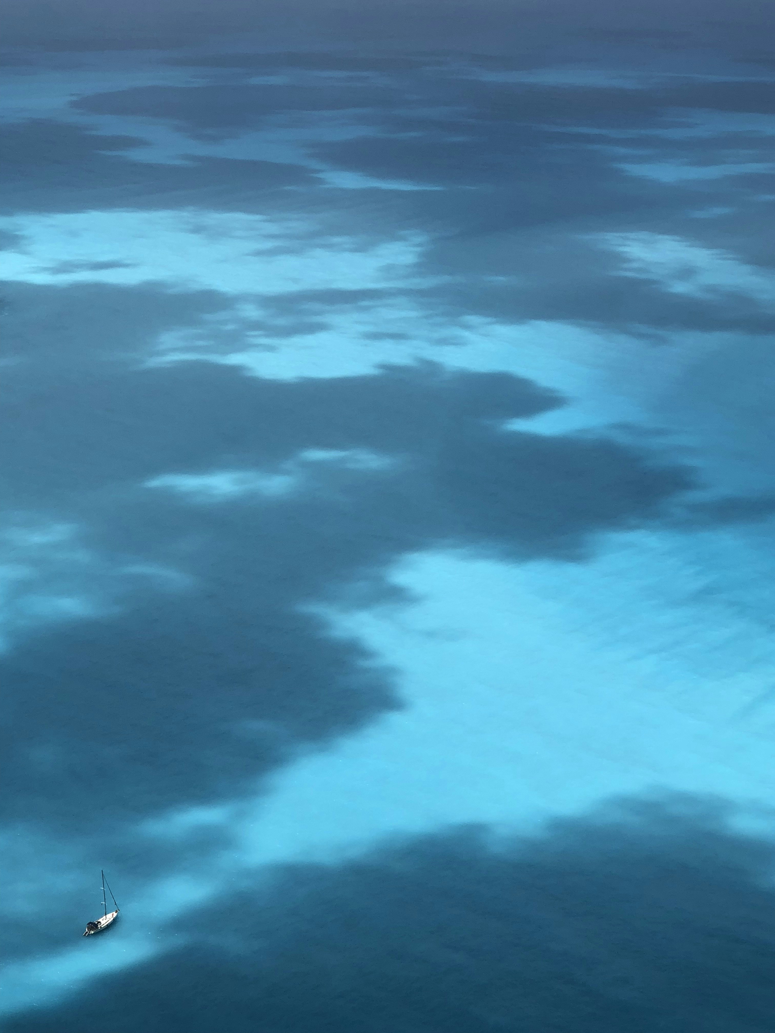 Lonely sailboat drifting across a vast expanse of turquoise water, surrounded by soft shadows and light patterns. 