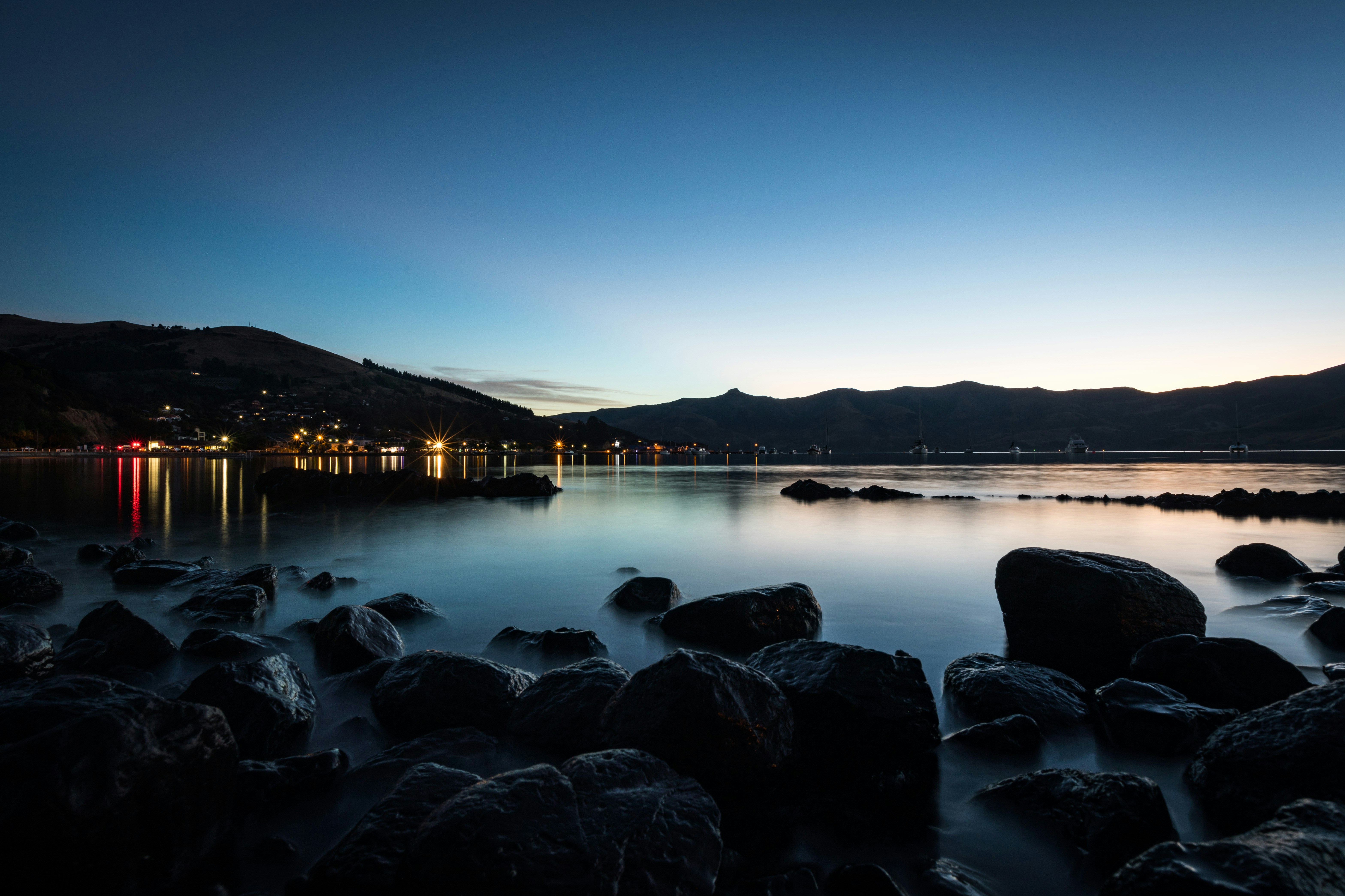 Smooth water surface reflecting distant lights and mountains at twilight, with dark rocks in the foreground.