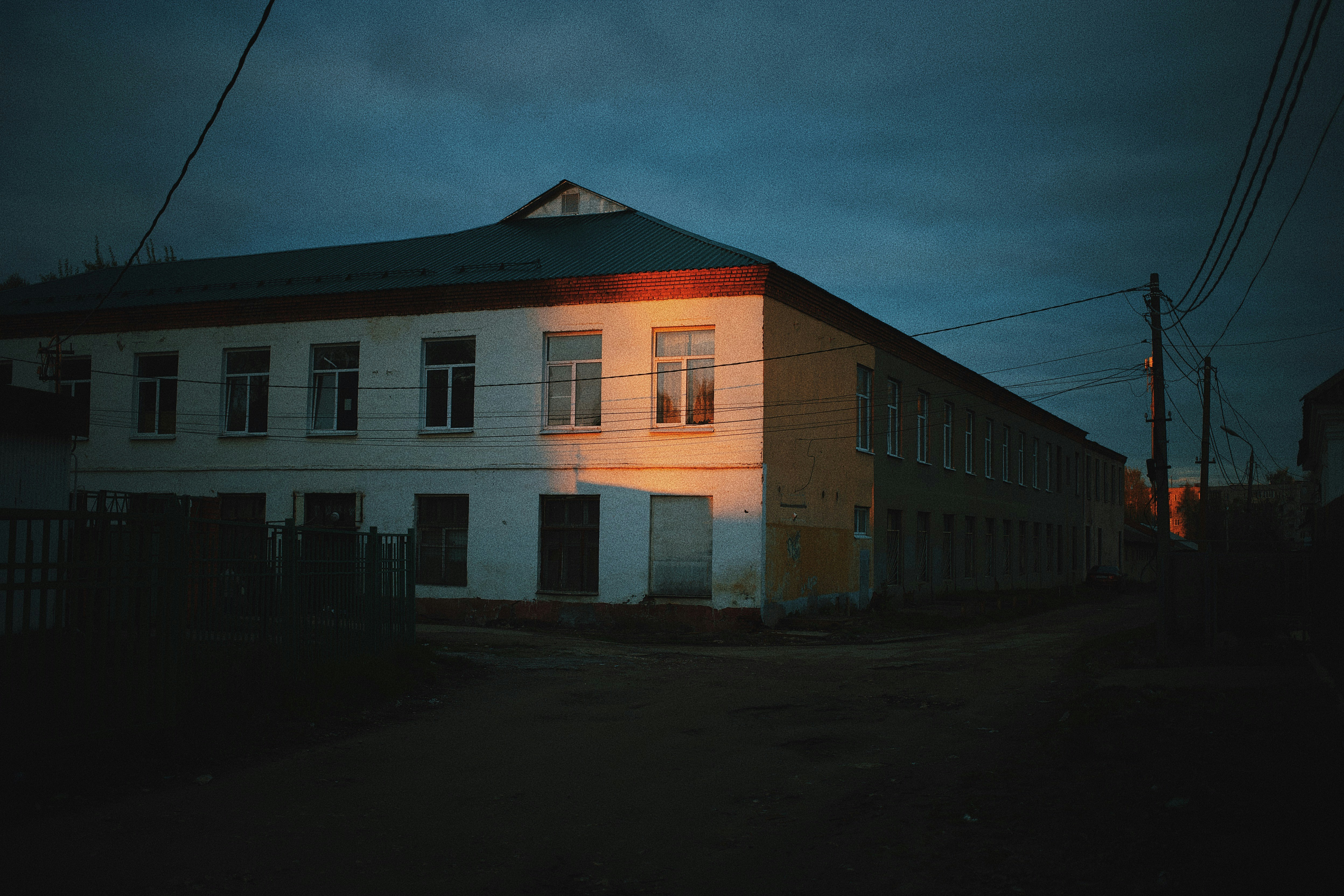 Two-story brick building bathed in blue hour light, with a warm orange glow highlighting the left façade; a quiet, desolate street scene.