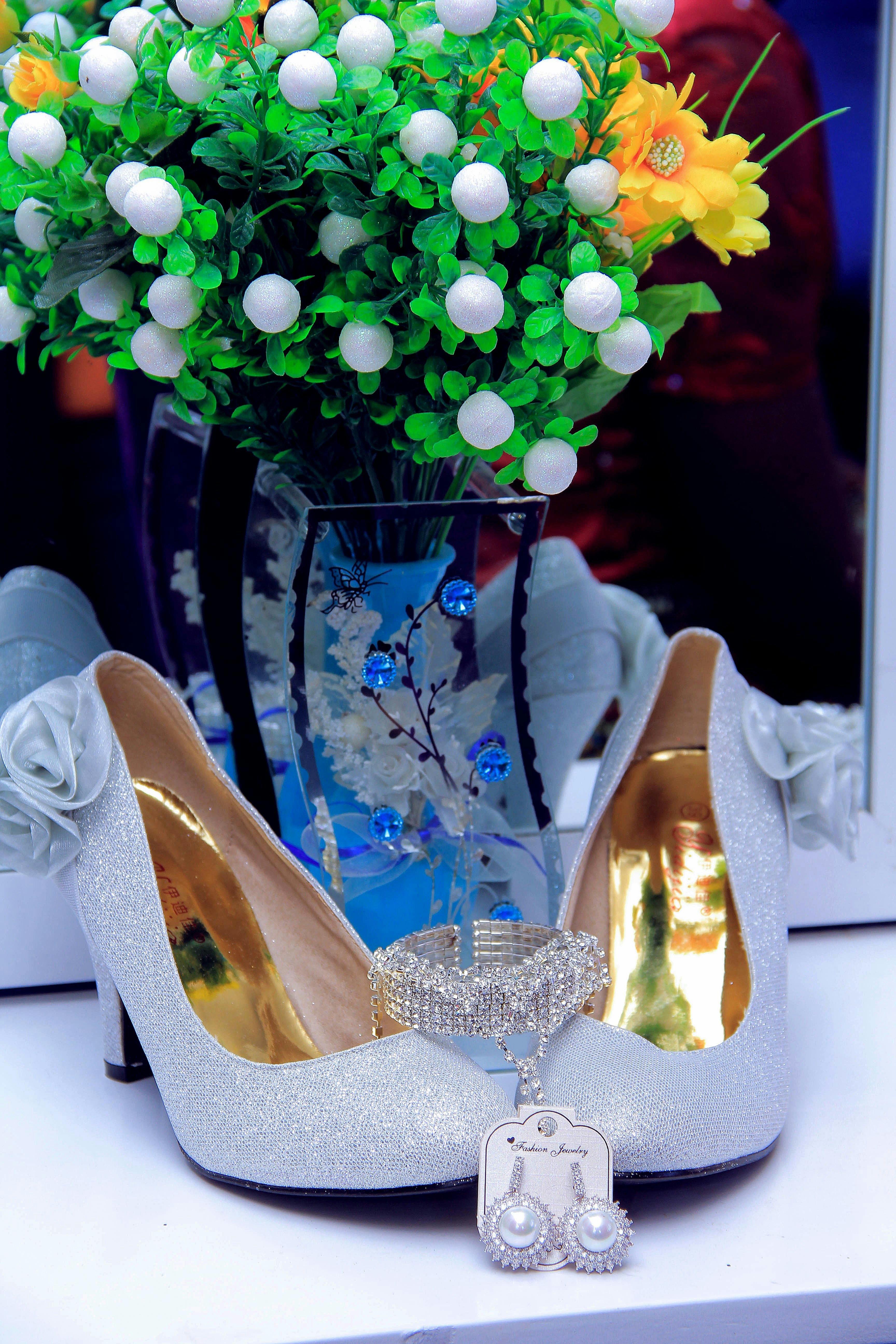 A pair of sparkling silver bridal shoes adorned with floral accents, accompanied by a delicate jewelry piece, set against a vibrant floral arrangement.