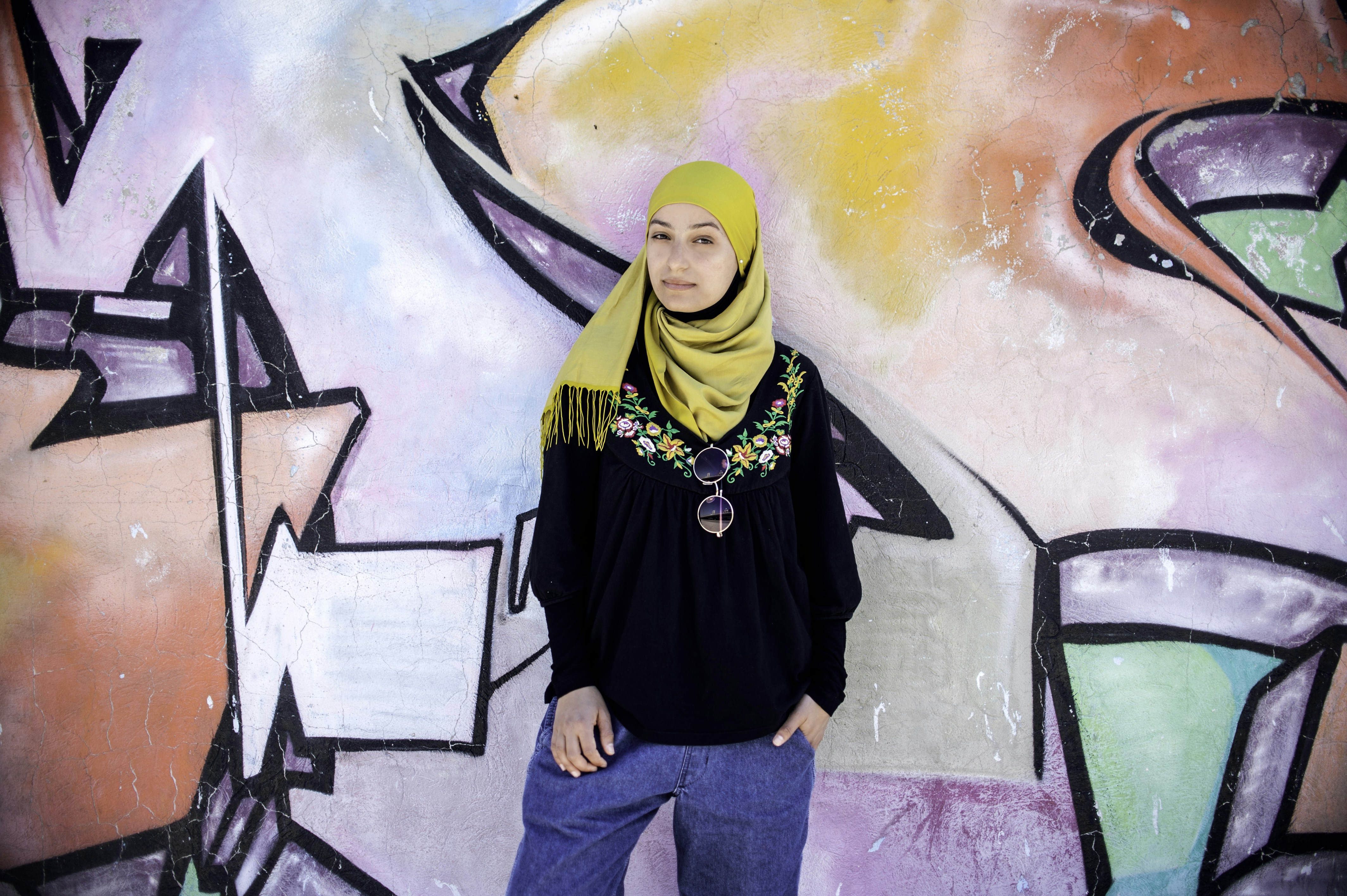 Young woman in a colorful hijab stands confidently before a graffiti mural, showcasing a blend of tradition and urban art.