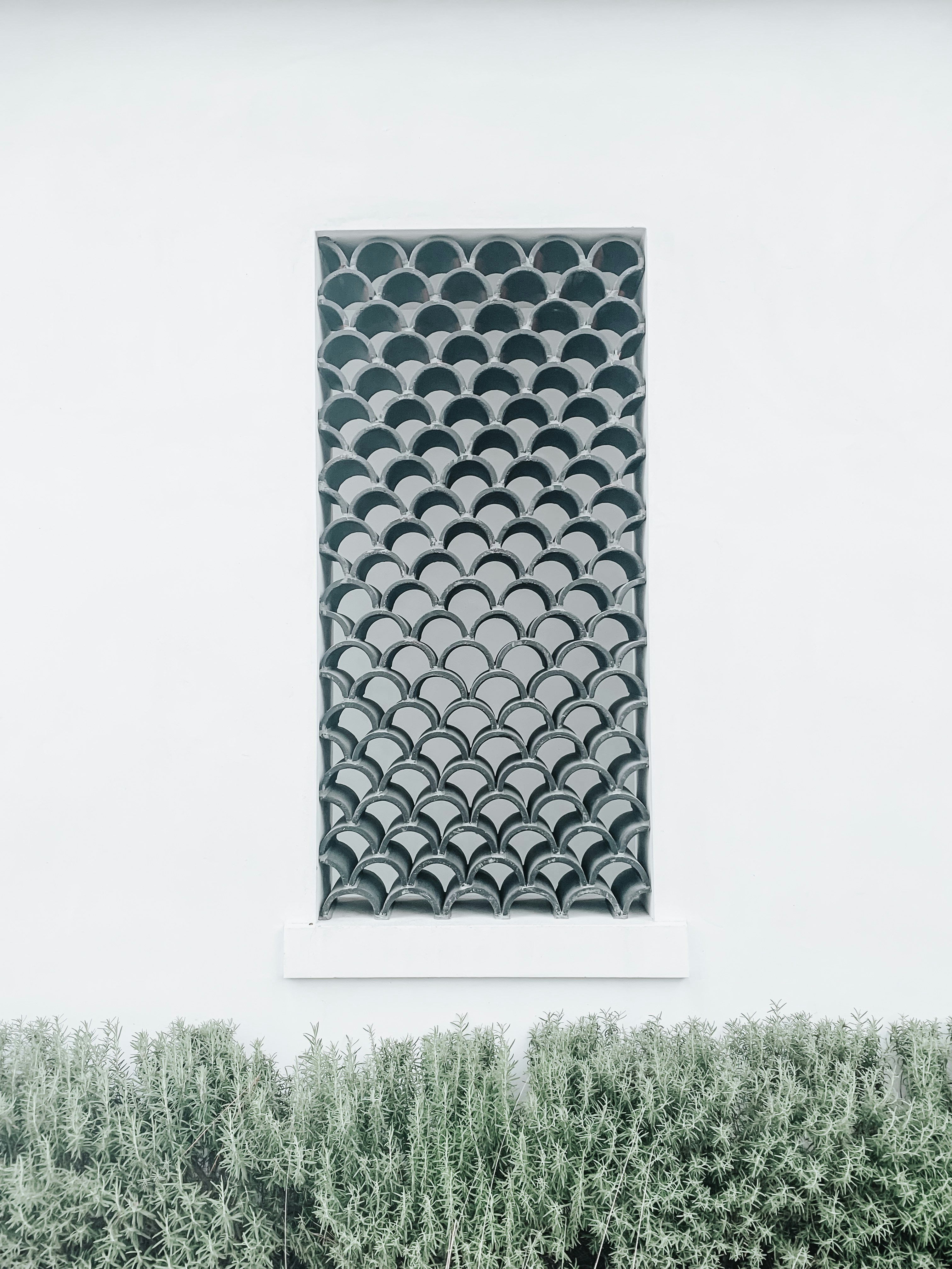 Intricate patterned wall feature with a series of scalloped shapes, framed by lush greenery at the base. The minimalistic design contrasts with the surrounding environment.