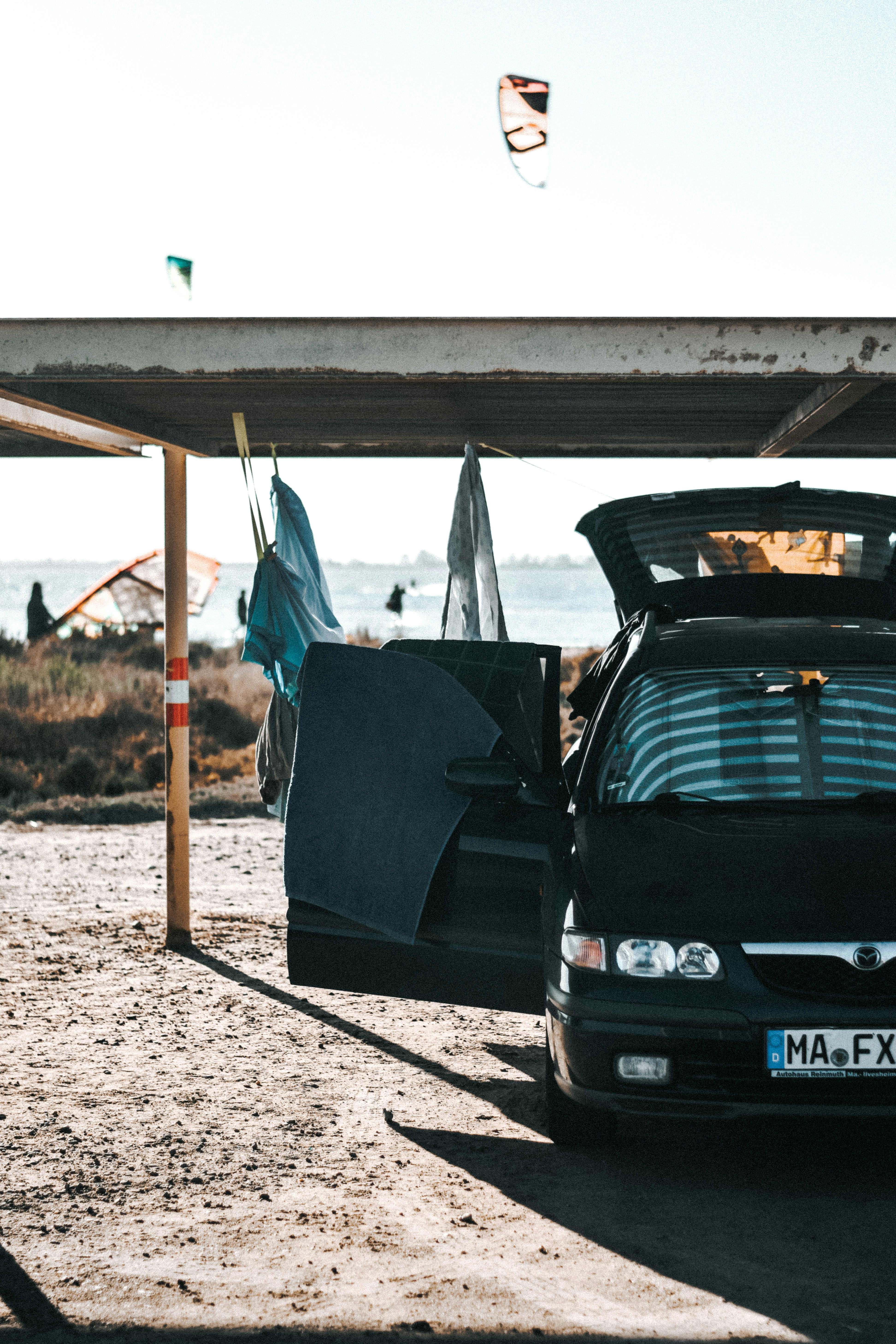 A black car parked under a shelter, with windsurfing gear hanging nearby, set against a backdrop of a sunny beach and colorful kites in the sky.