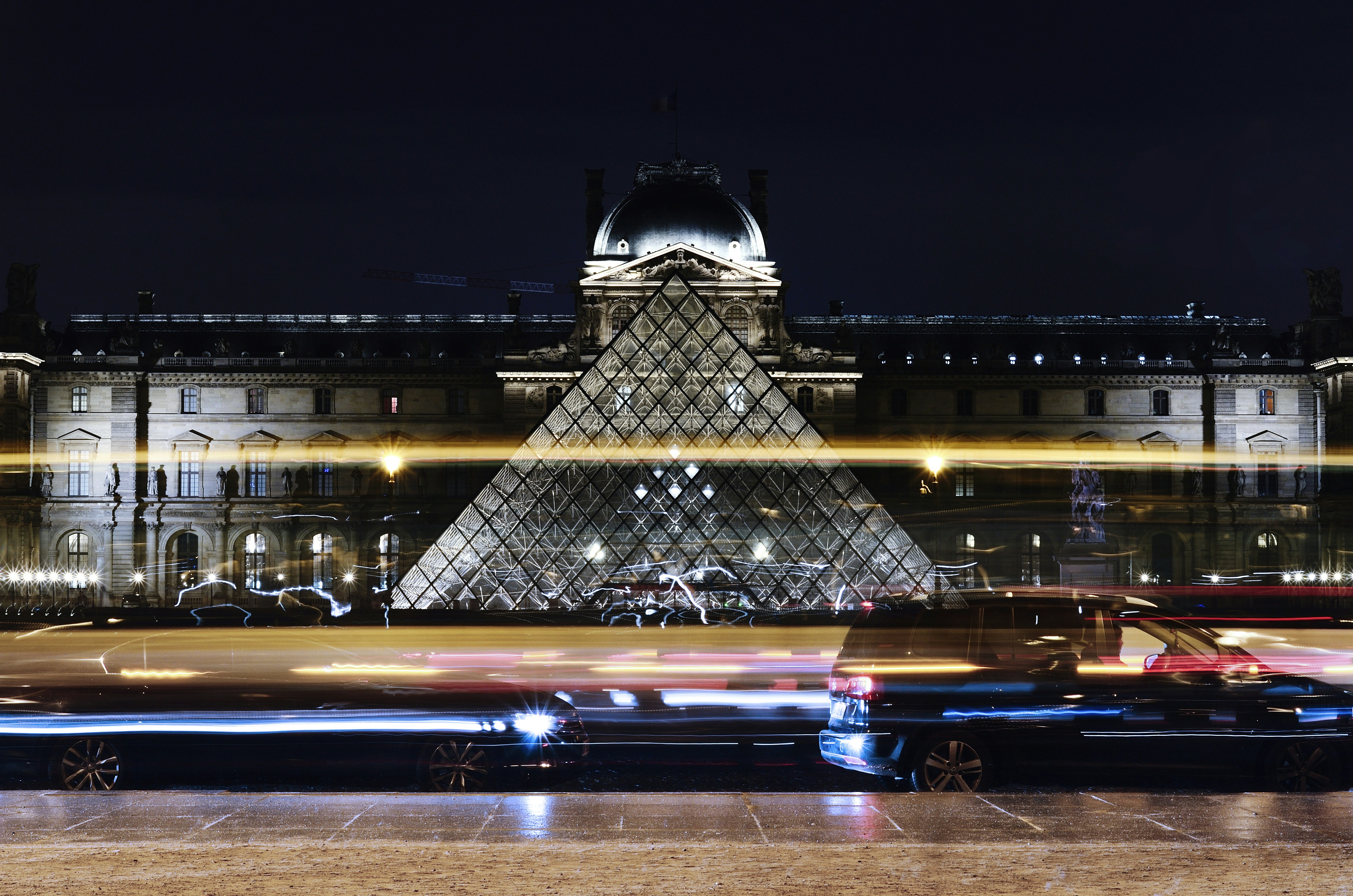 The iconic glass pyramid of the Louvre Museum glows against a backdrop of historical architecture, with streaks of light from passing vehicles creating a dynamic contrast.