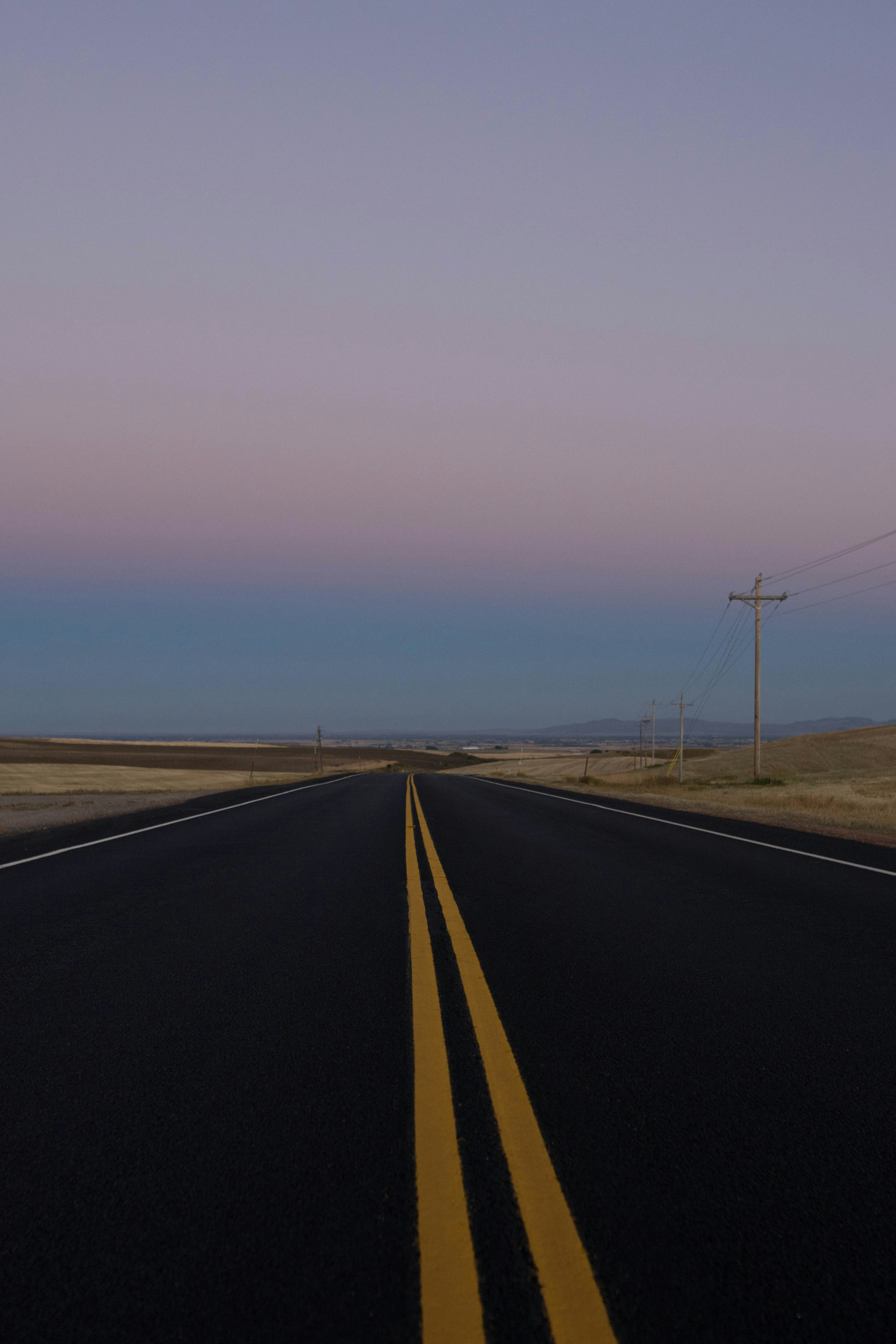 A long, straight road stretches into the distance, flanked by fields under a pastel sky at dusk.