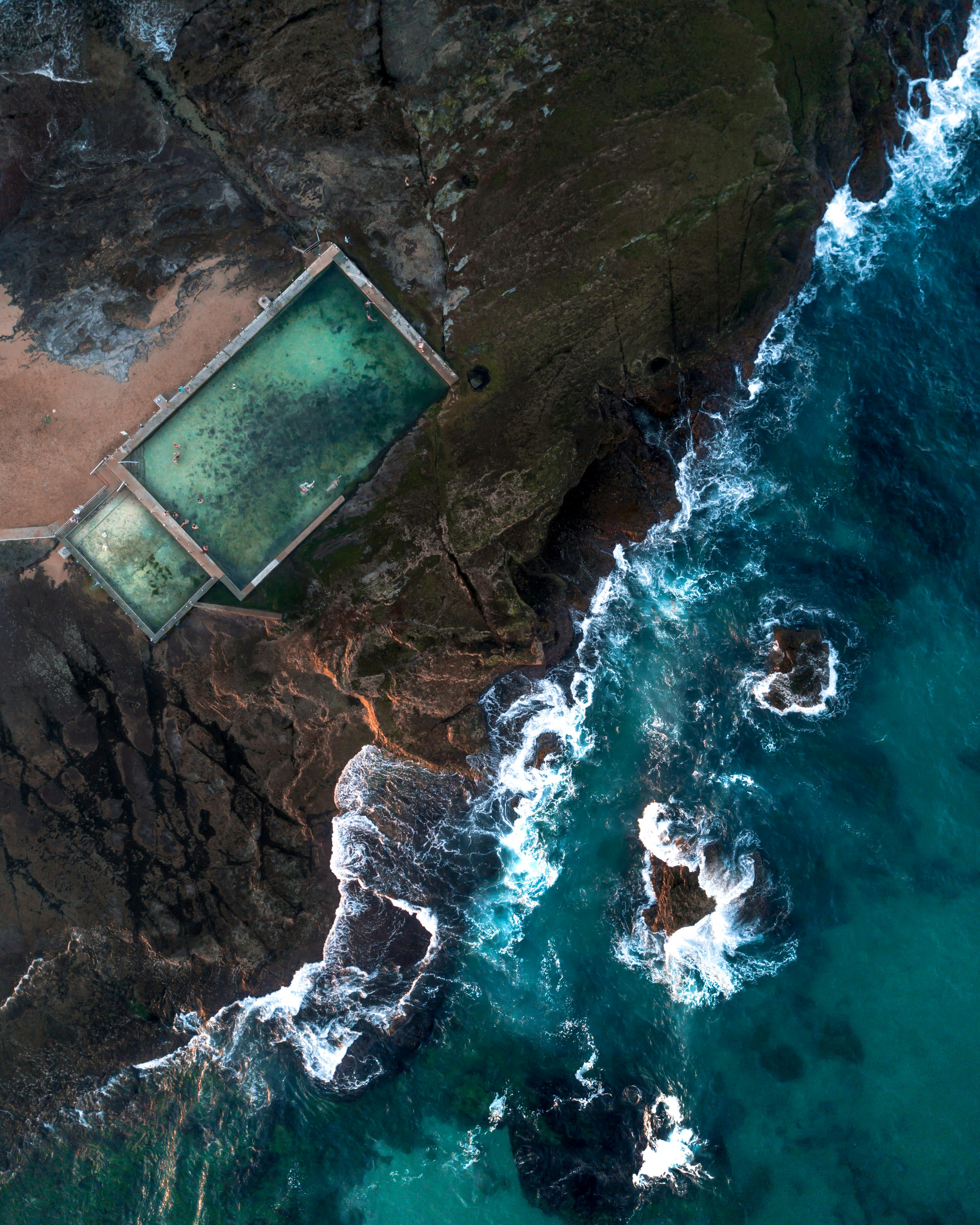 Aerial view of a coastal pool adjacent to rugged rocks and vibrant ocean waves.