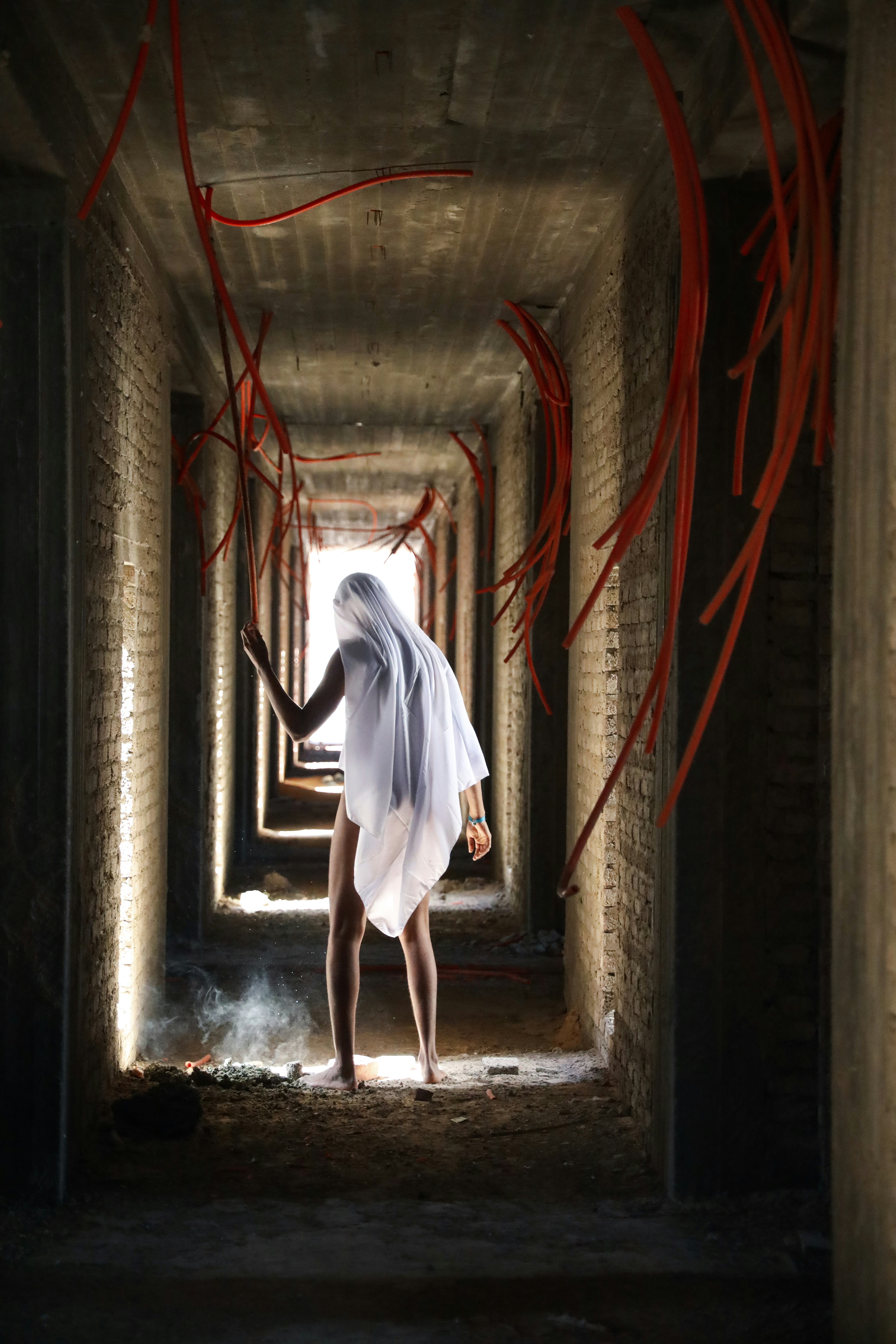 A figure draped in white stands in a dimly lit corridor, surrounded by exposed wiring and dust, evoking a sense of mystery and isolation.