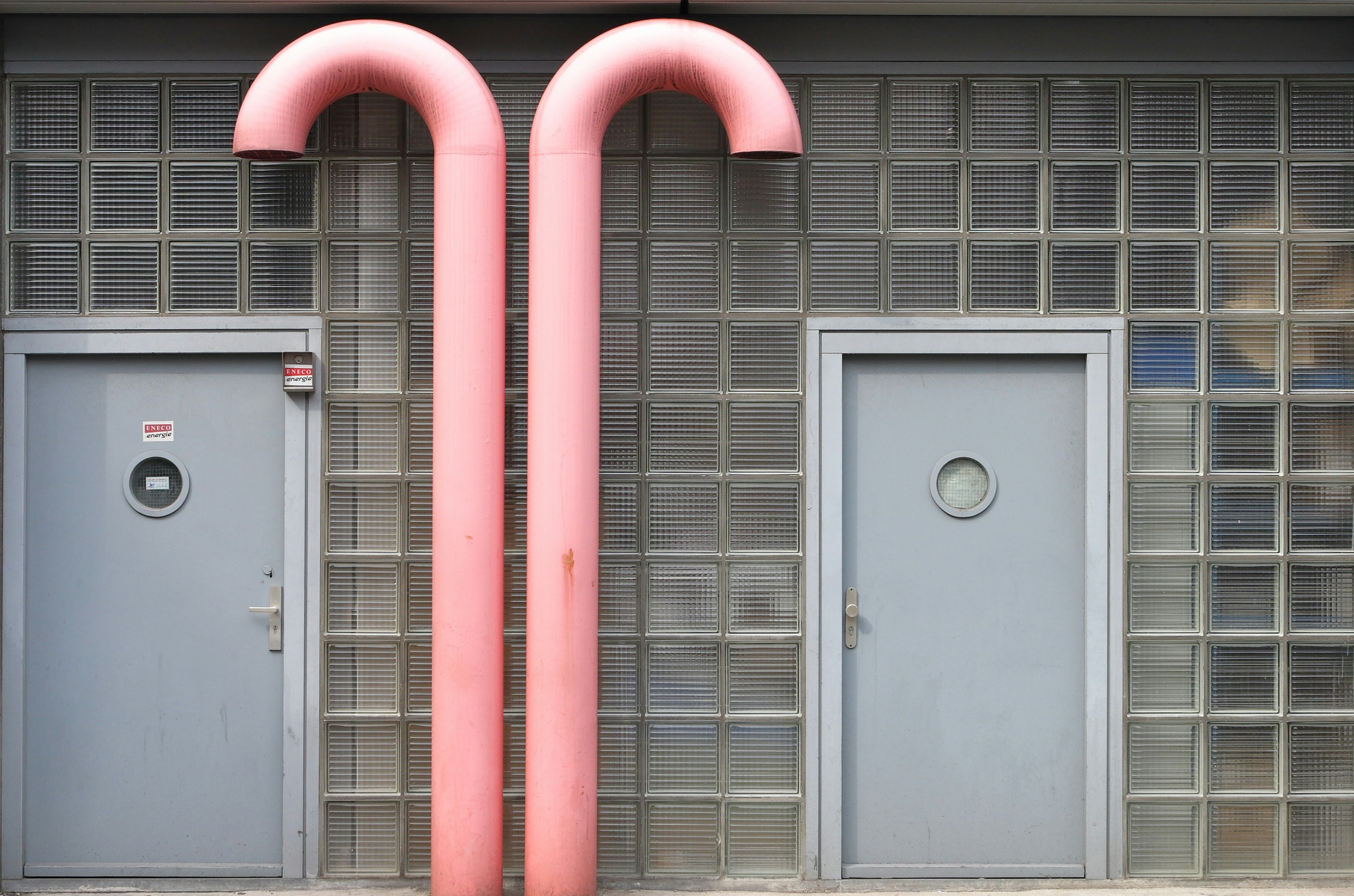 Two gray doors flank a pair of oversized pink pipes against a textured glass block wall, creating a striking contrast in a minimalist setting.