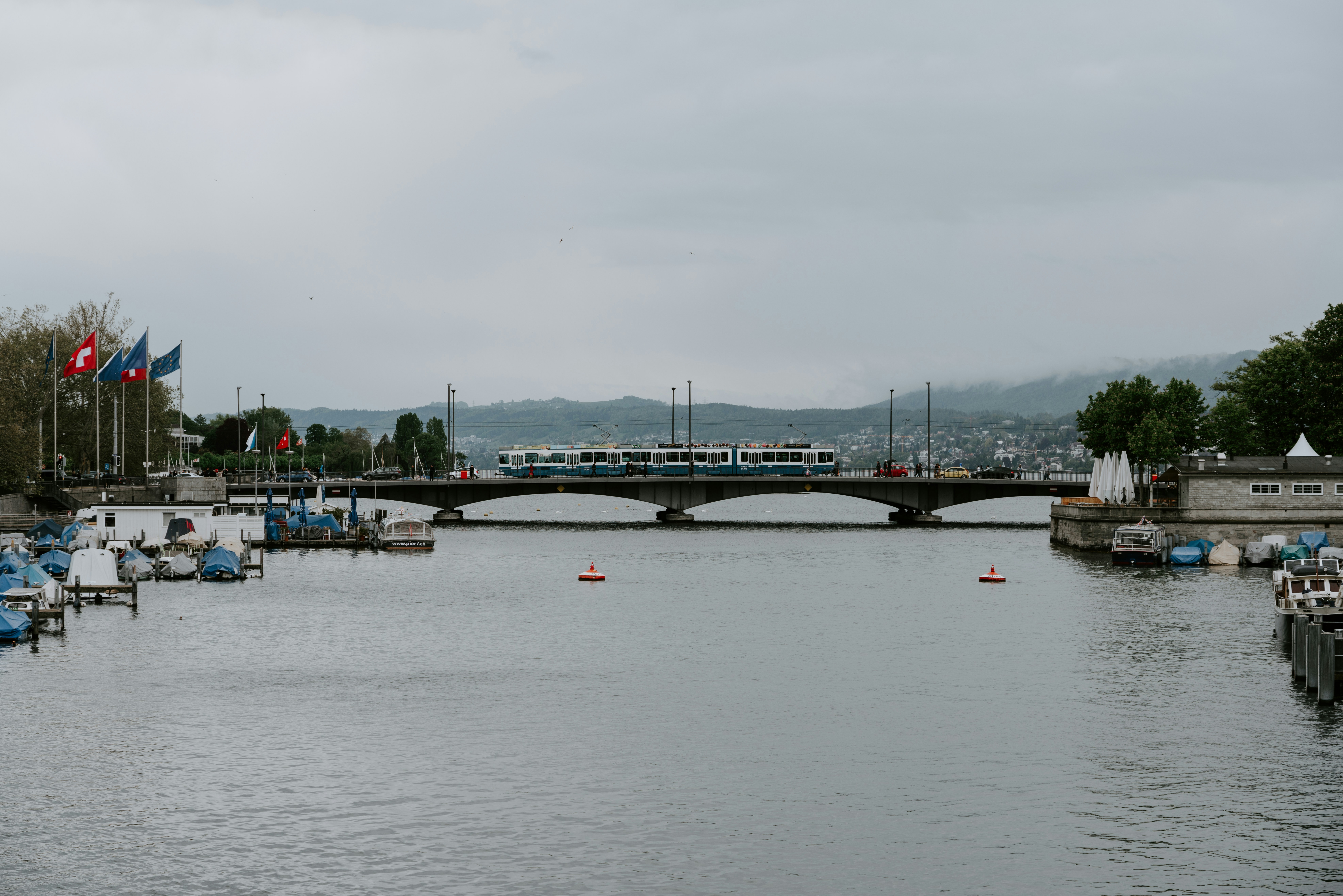 A serene view of a river with boats and a bridge in the background, framed by flags and greenery. The scene captures the calm essence of urban life by the water.