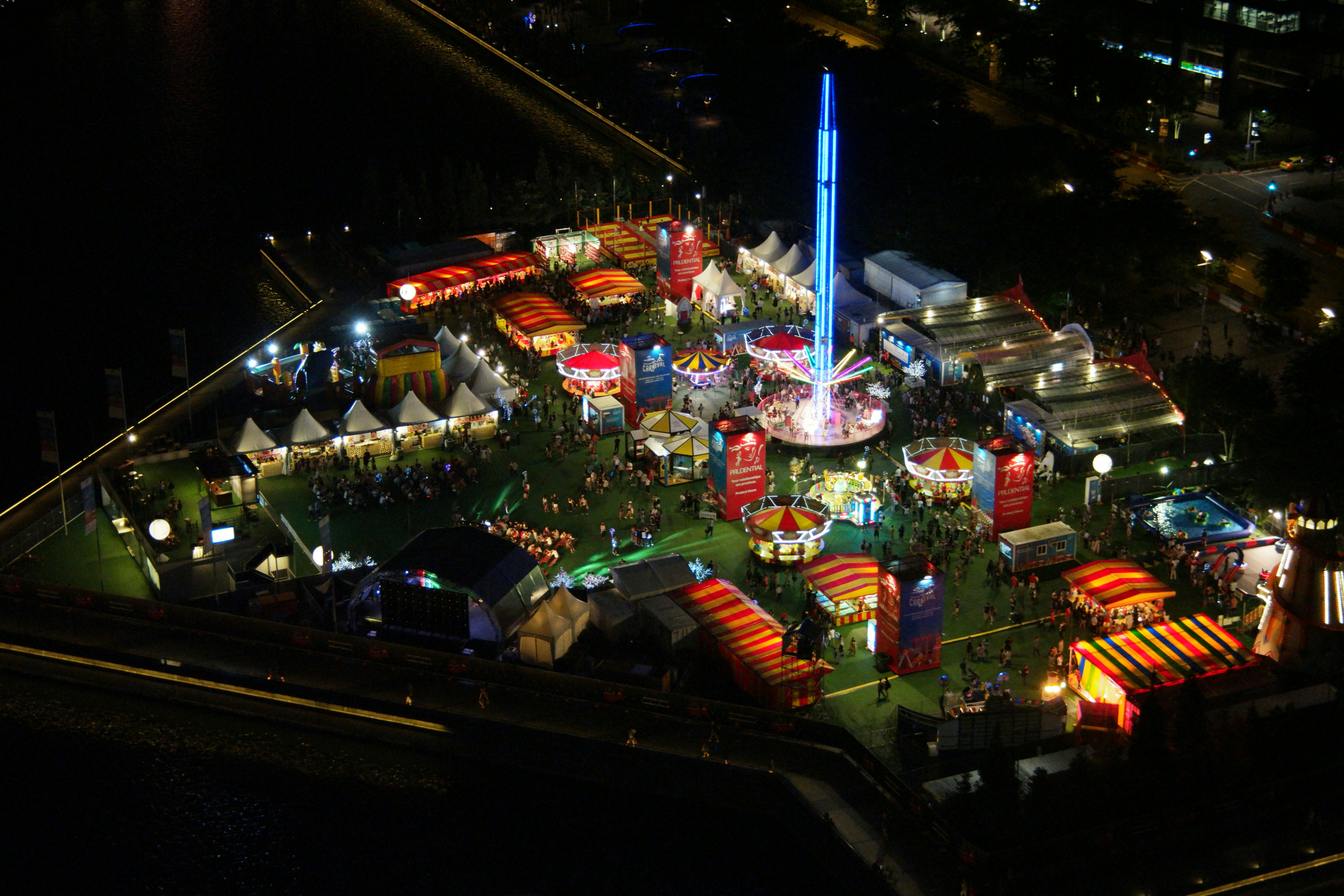 Aerial view of a lively carnival scene featuring colorful tents, attractions, and a central illuminated ride, bustling with visitors under the night sky.