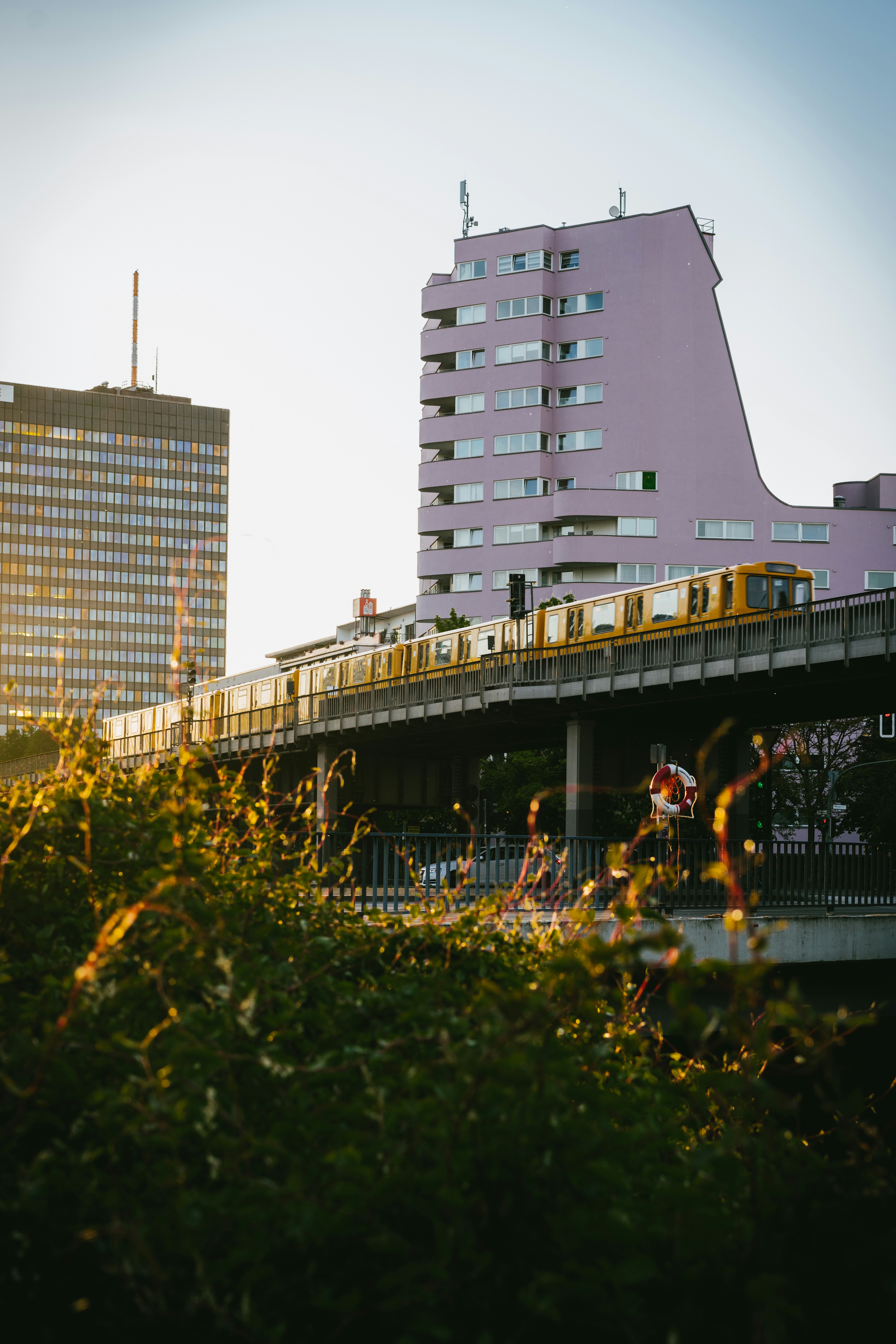 A vibrant scene showcasing a train crossing a bridge with modern architecture in the background, framed by lush greenery below. The warm light of sunset enhances the urban landscape.