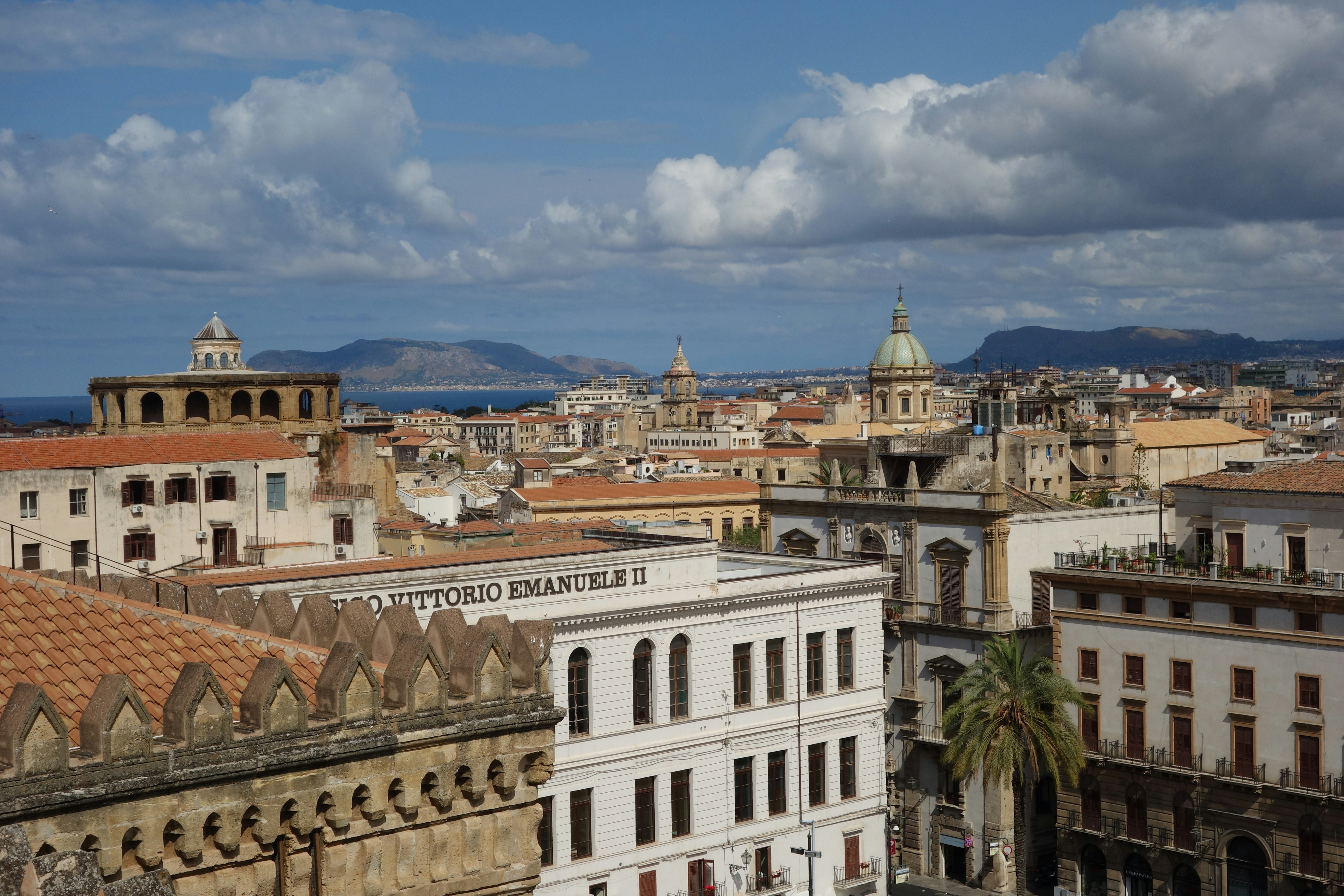 Panoramic view of a historic cityscape with terracotta rooftops and distant mountains under a partly cloudy sky.