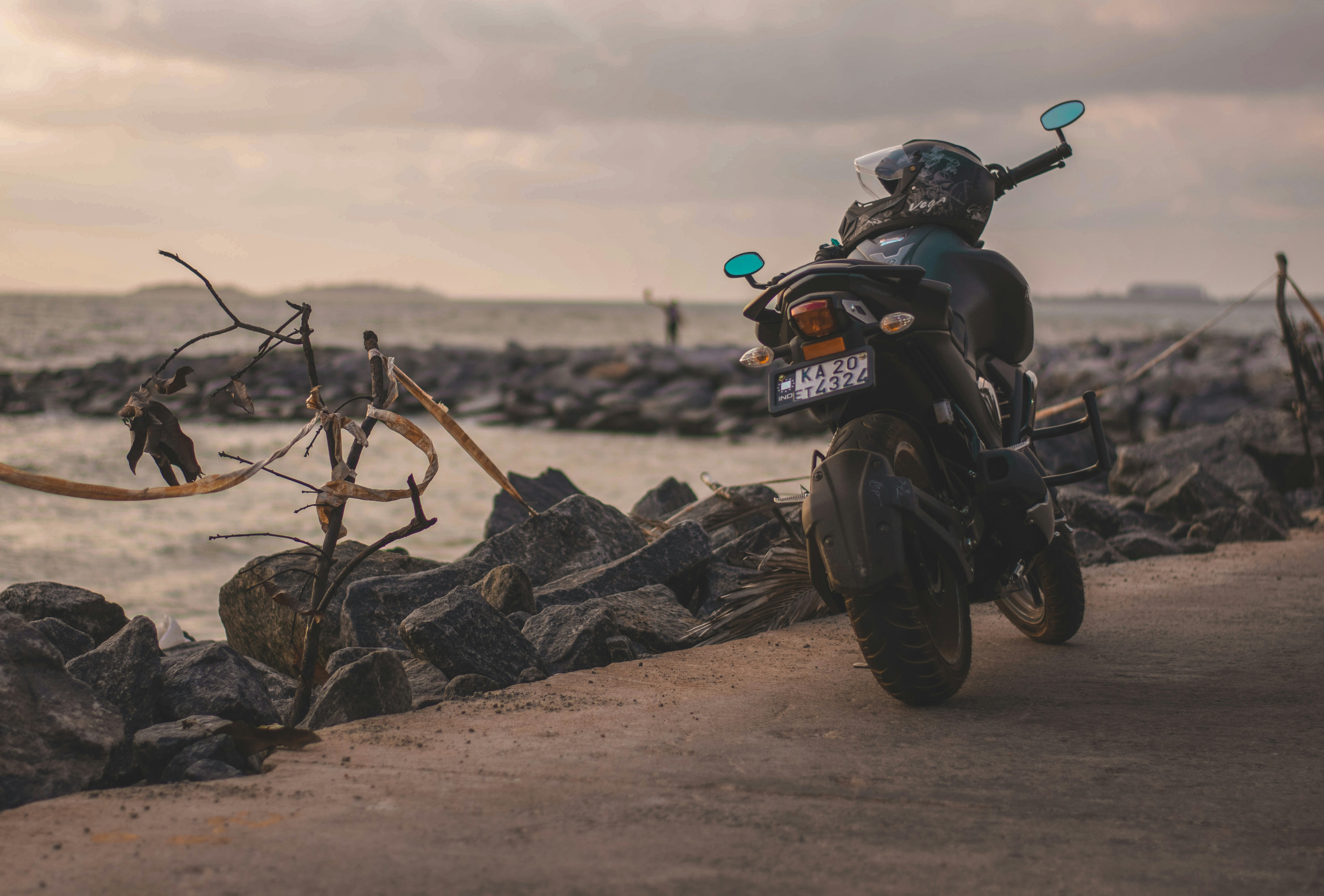 Motorcycle parked by rocky shoreline with distant figures and a cloudy sky. The scene evokes a sense of adventure and tranquility.