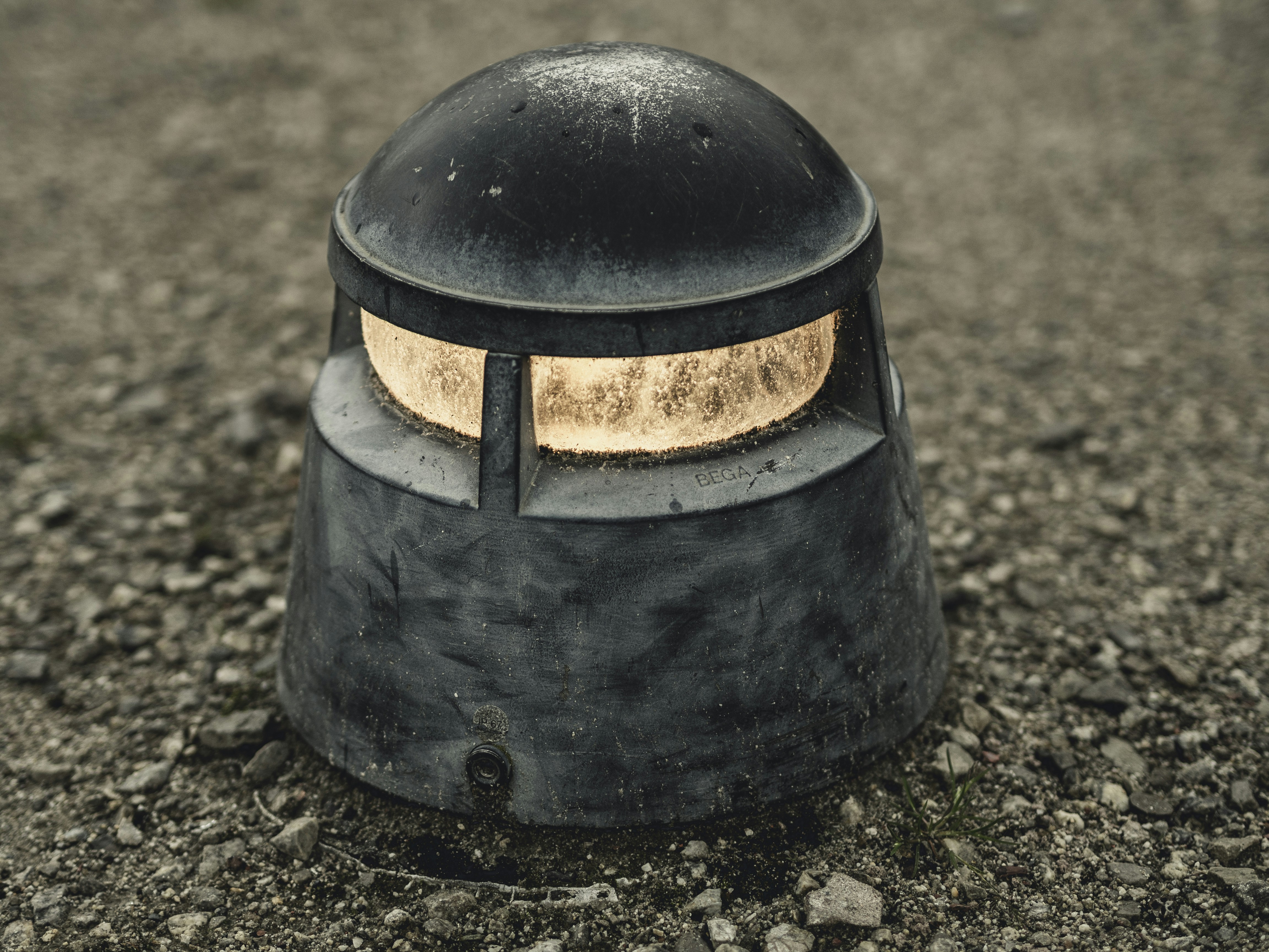 Weathered ground light fixture casting a soft glow on gravel surface, highlighting its rugged texture and design.