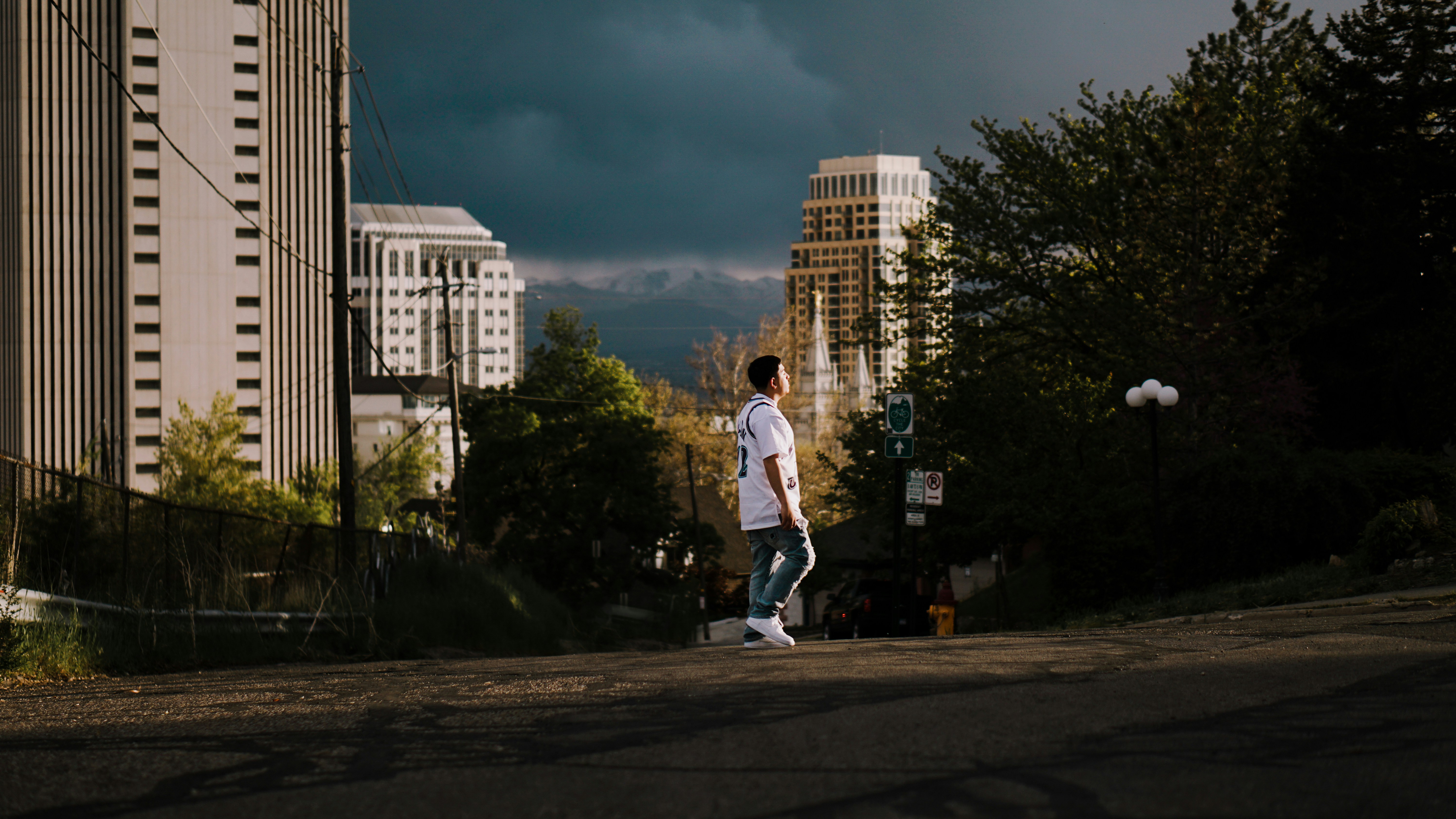 Person standing on a sunlit path between towering city buildings under a dramatic sky.