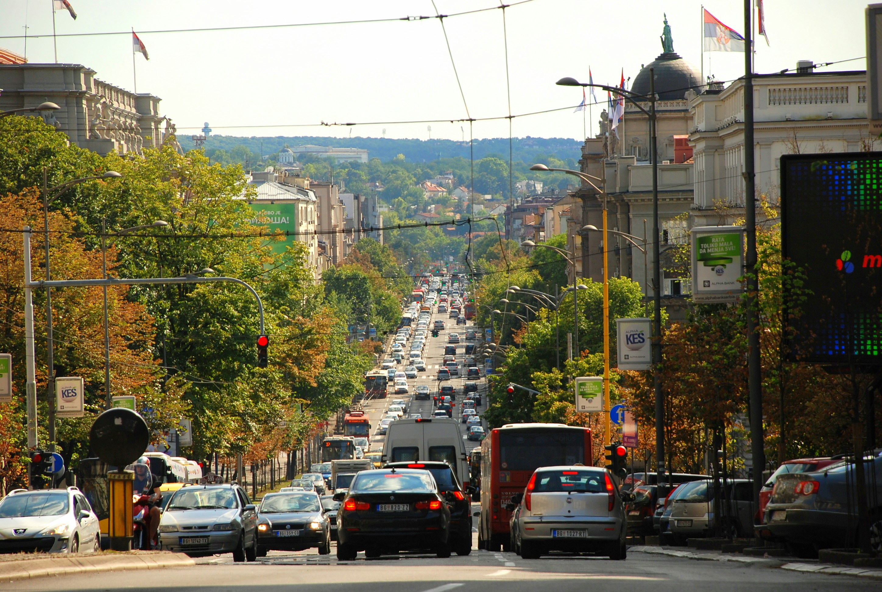 Colorful cars navigate a bustling city street lined with trees and historic buildings.