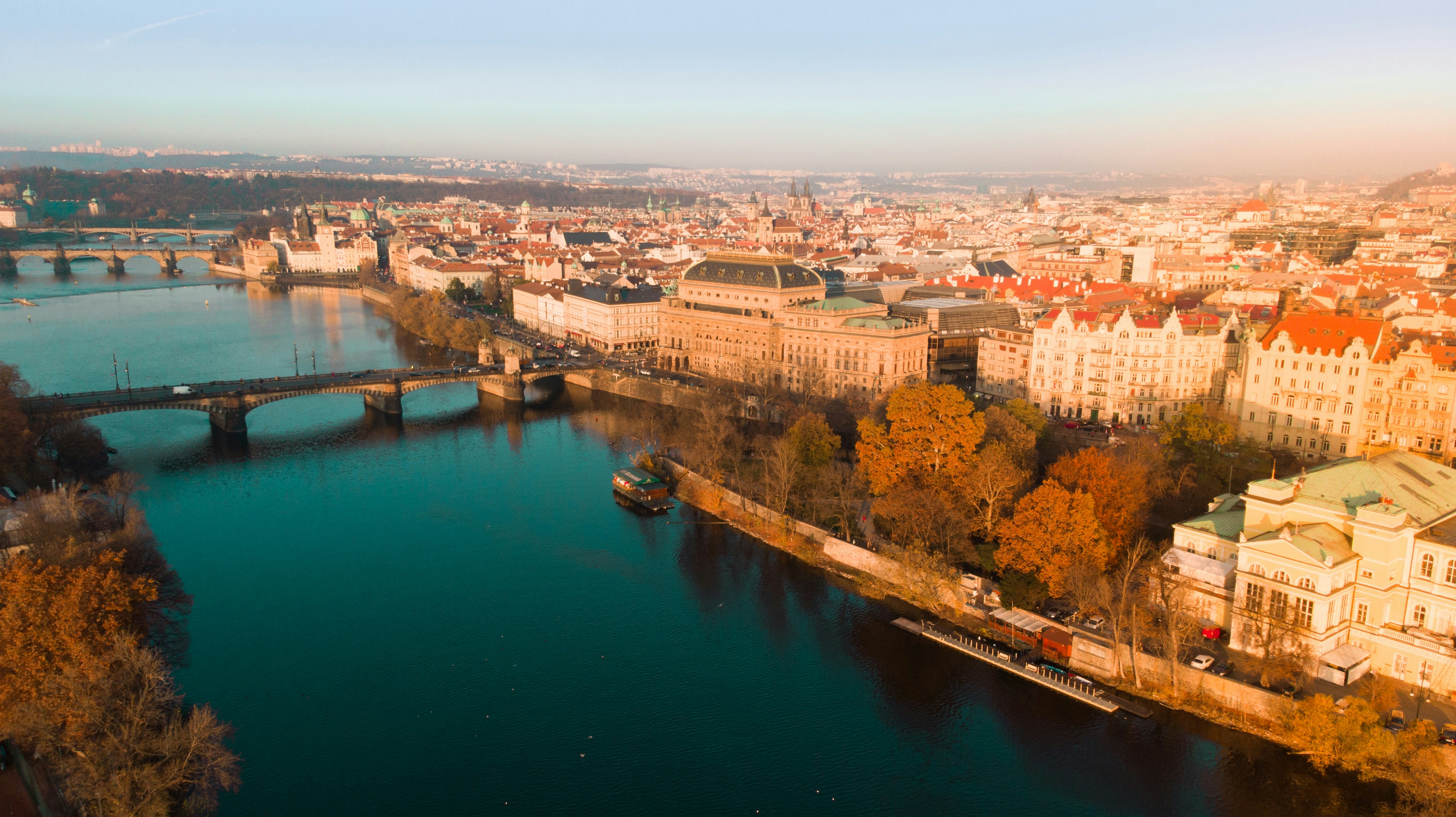 Aerial view of a city with historic bridges spanning a wide river, framed by autumn foliage and bathed in warm sunlight.