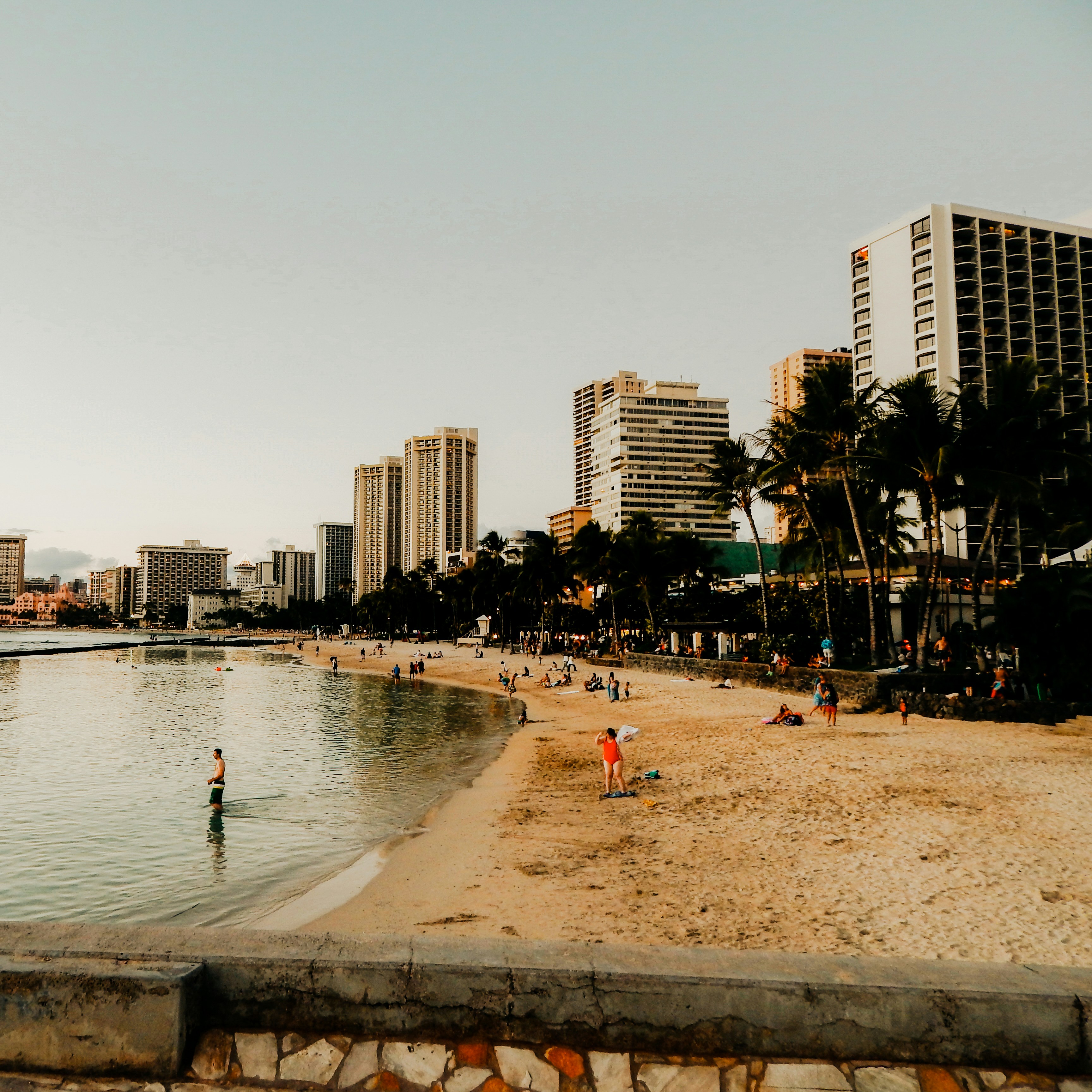 Crowded beach scene at sunset with city skyline in the background, showcasing people enjoying the shoreline and warm hues of the evening sky.