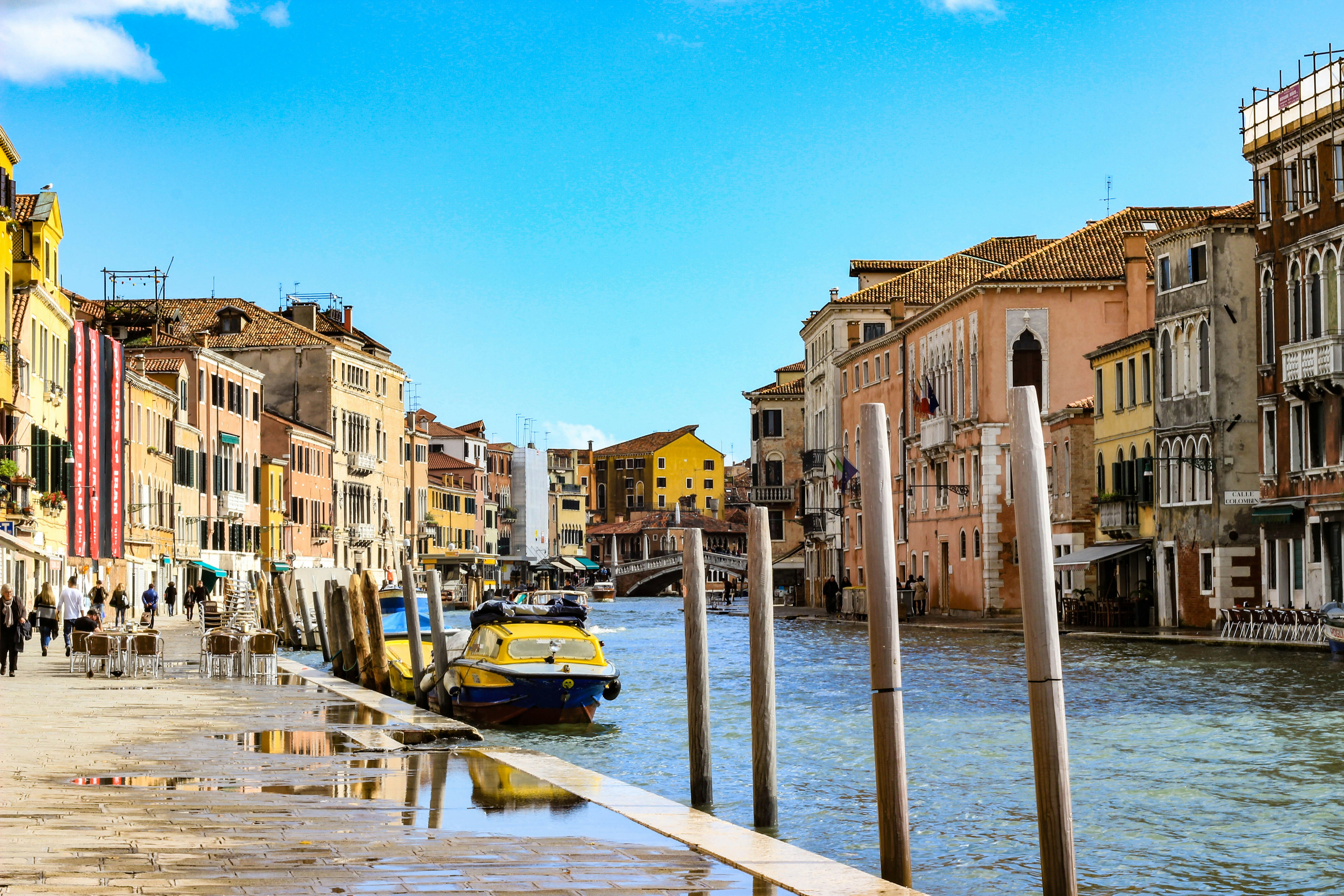 This vibrant image captures a picturesque Venetian canal, flanked by colorful, historic buildings under a bright blue sky. The composition leads the eye along the waterway, with reflections shimmering on the wet pavement, adding depth and interest. The lively hues of yellow and pink buildings create a warm, inviting atmosphere, making the scene visually striking and evocative of Venice's unique charm.