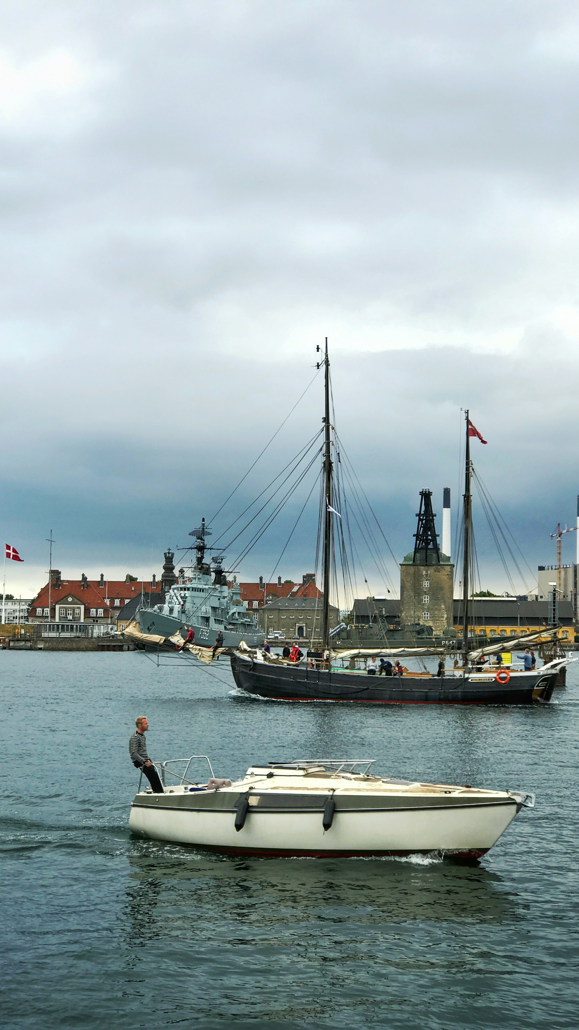 A small white boat navigates through a bustling harbor, surrounded by historic vessels and charming waterfront buildings. The cloudy sky adds a dramatic backdrop.