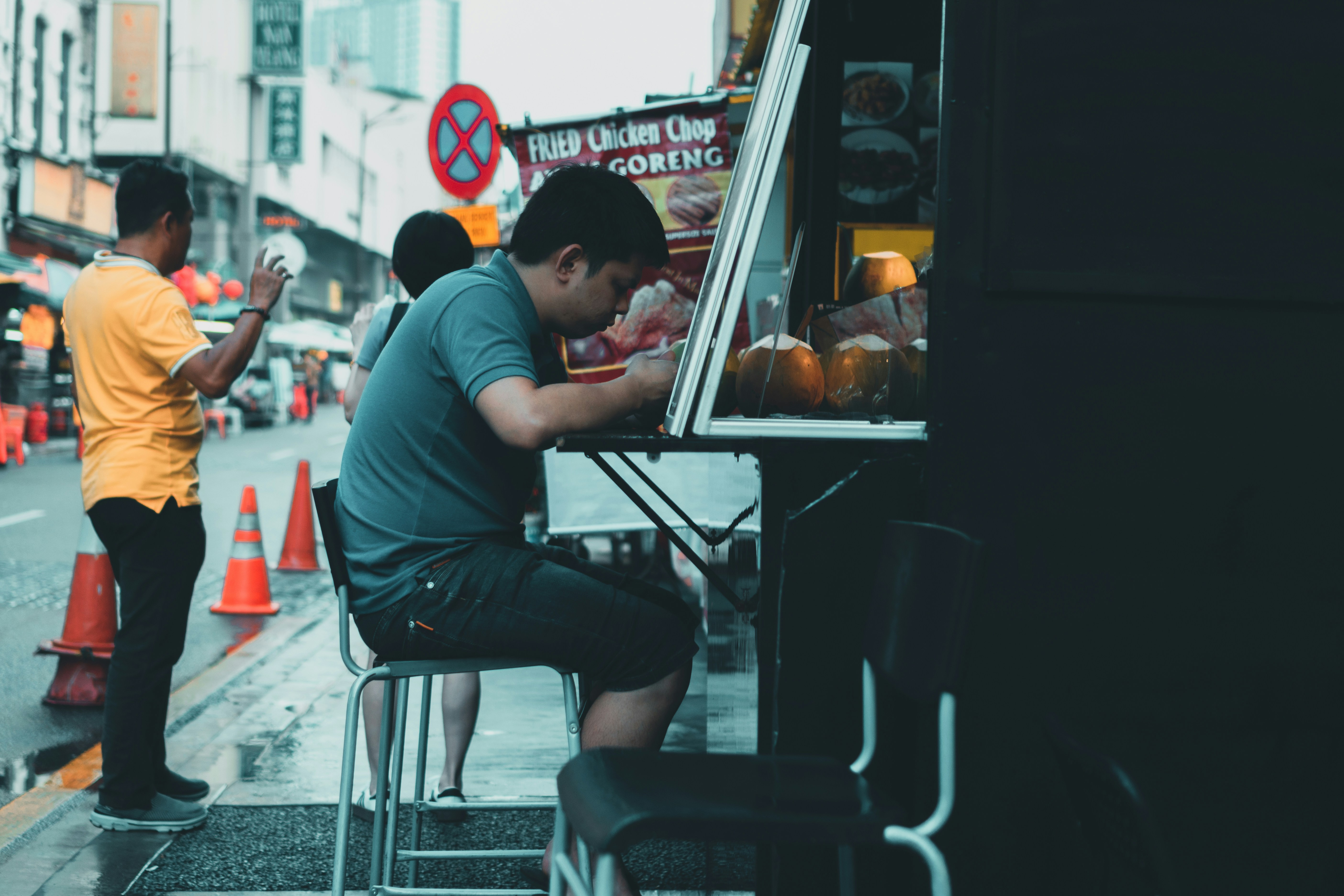A young man seated at a food stall, deeply focused on his meal, while the vibrant street scene unfolds around him. The setting captures the essence of urban dining.
