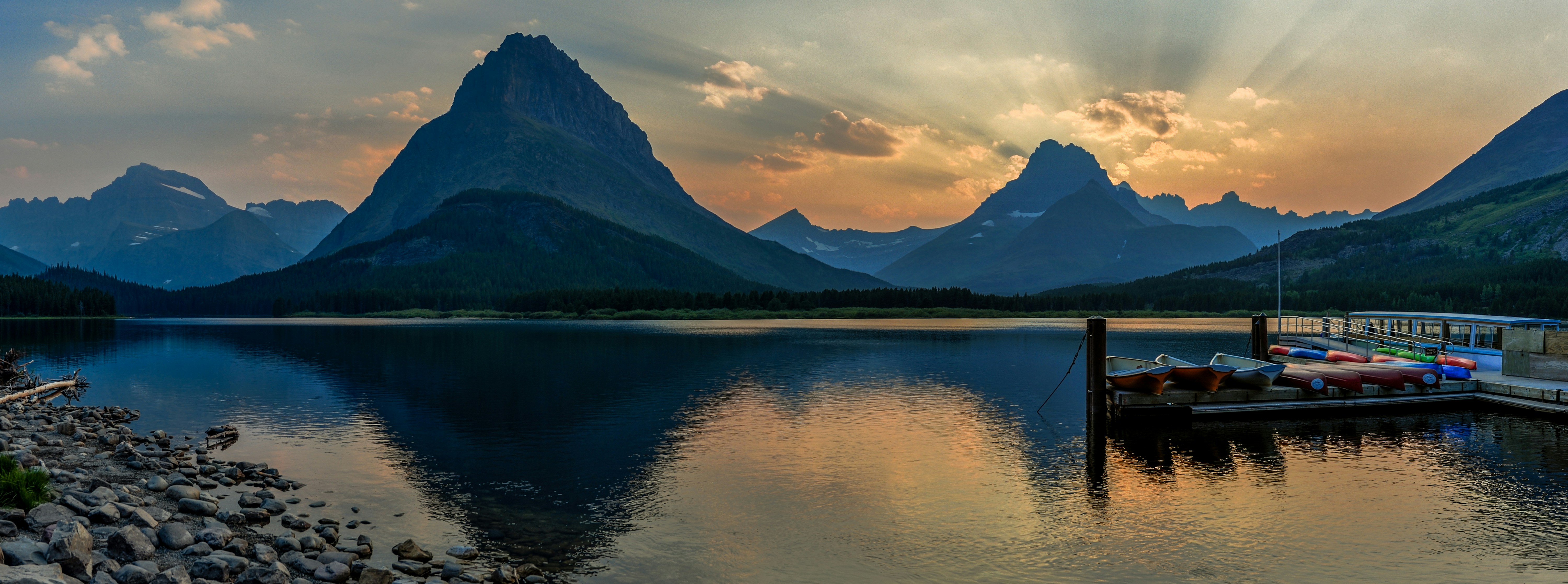 Serene lake mirroring jagged mountain peaks under a dramatic sunset sky.