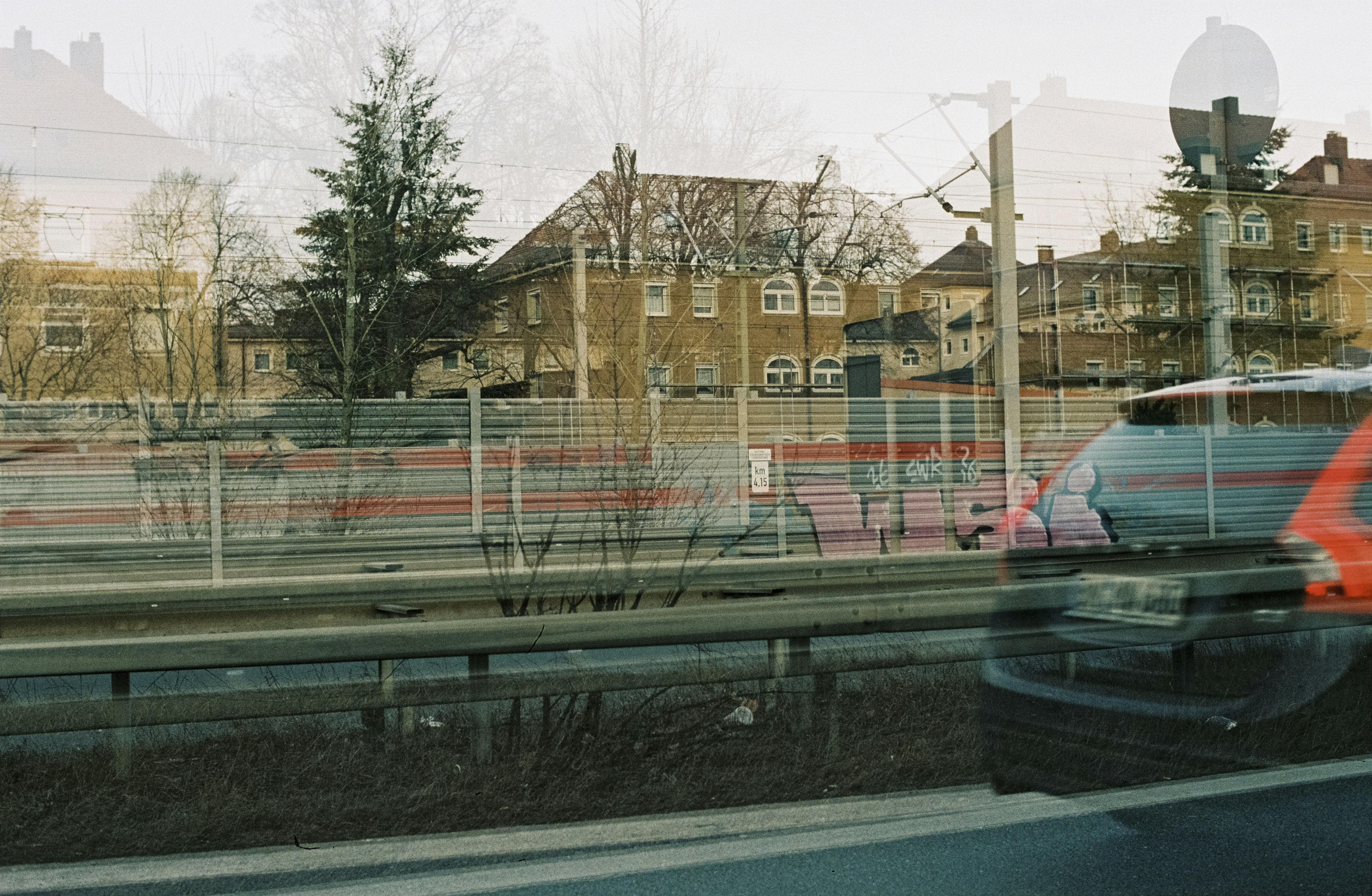 A multilayer urban street photograph with a moving blue car superimposed over a row of brick houses and a guardrail, creating a double-exposure effect. The composition emphasizes motion and reflections on a glass surface.